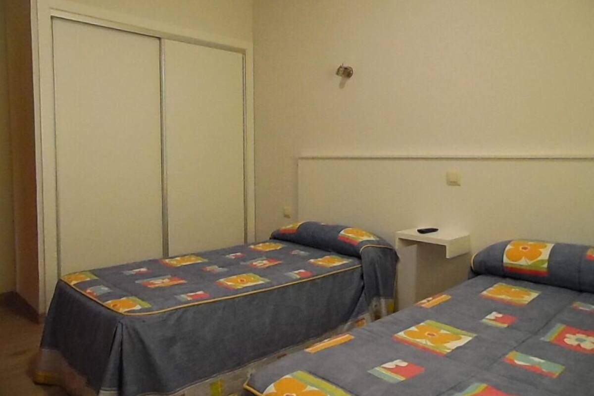 Quarto com duas camas, cada uma coberta por um edredom azul com padrões coloridos. Há uma mesa de cabeceira branca com um controle remoto em cima.