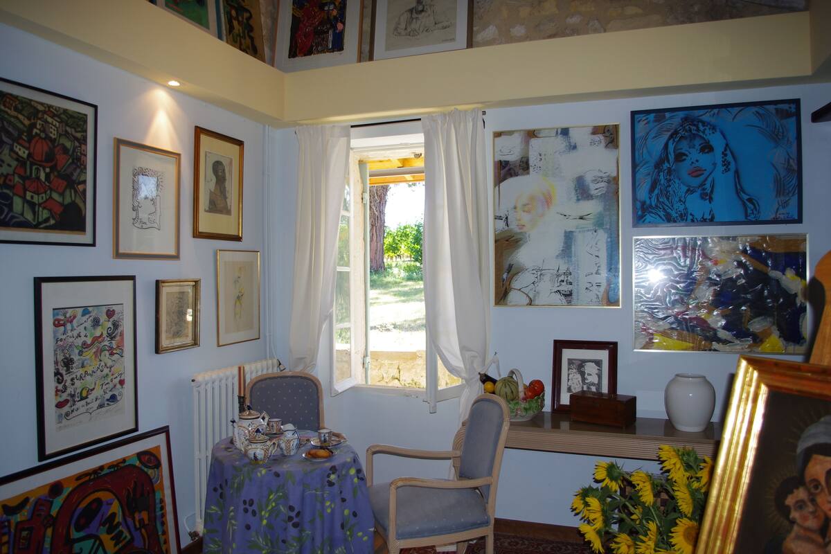 Quarto com uma mesa de jantar, uma cadeira, janela com cortinas e diversas obras de arte emolduradas nas paredes.