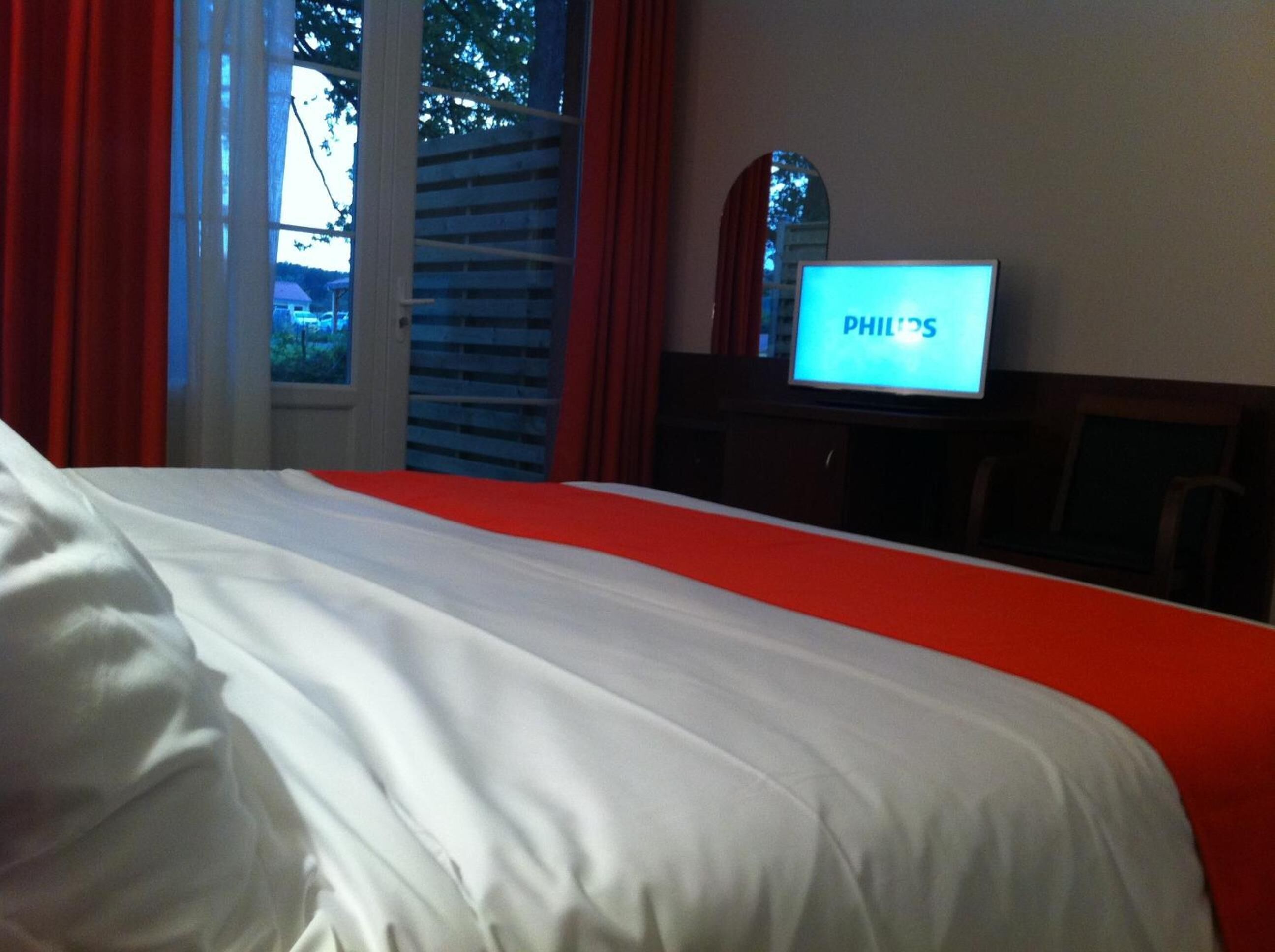 Quarto de hotel com uma cama, uma televisão exibindo "PHILIPS" na tela, e vista do exterior através de uma janela com cortinas vermelhas.
