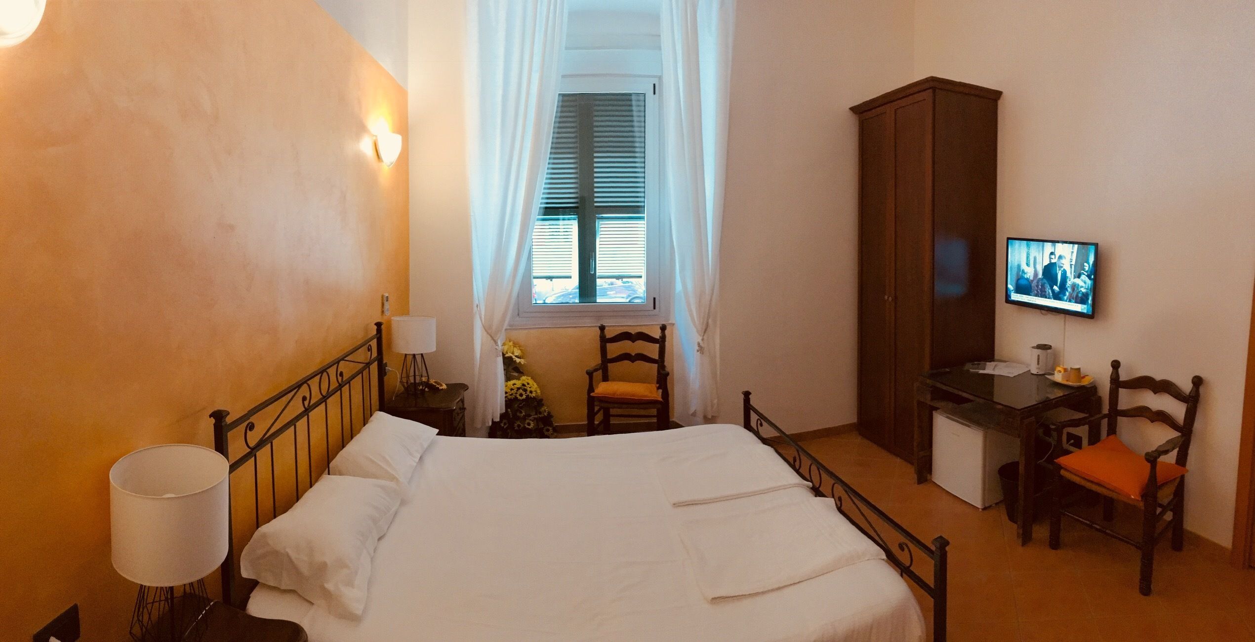 Quarto com cama, escrivaninha, cadeira, televisão e janela com cortinas.