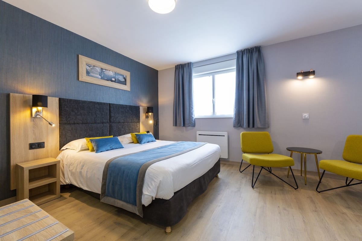 Quarto de hotel moderno com uma cama grande, duas mesas de cabeceira, uma mesinha, duas cadeiras amarelas, uma janela com cortinas e um quadro fixado na parede.