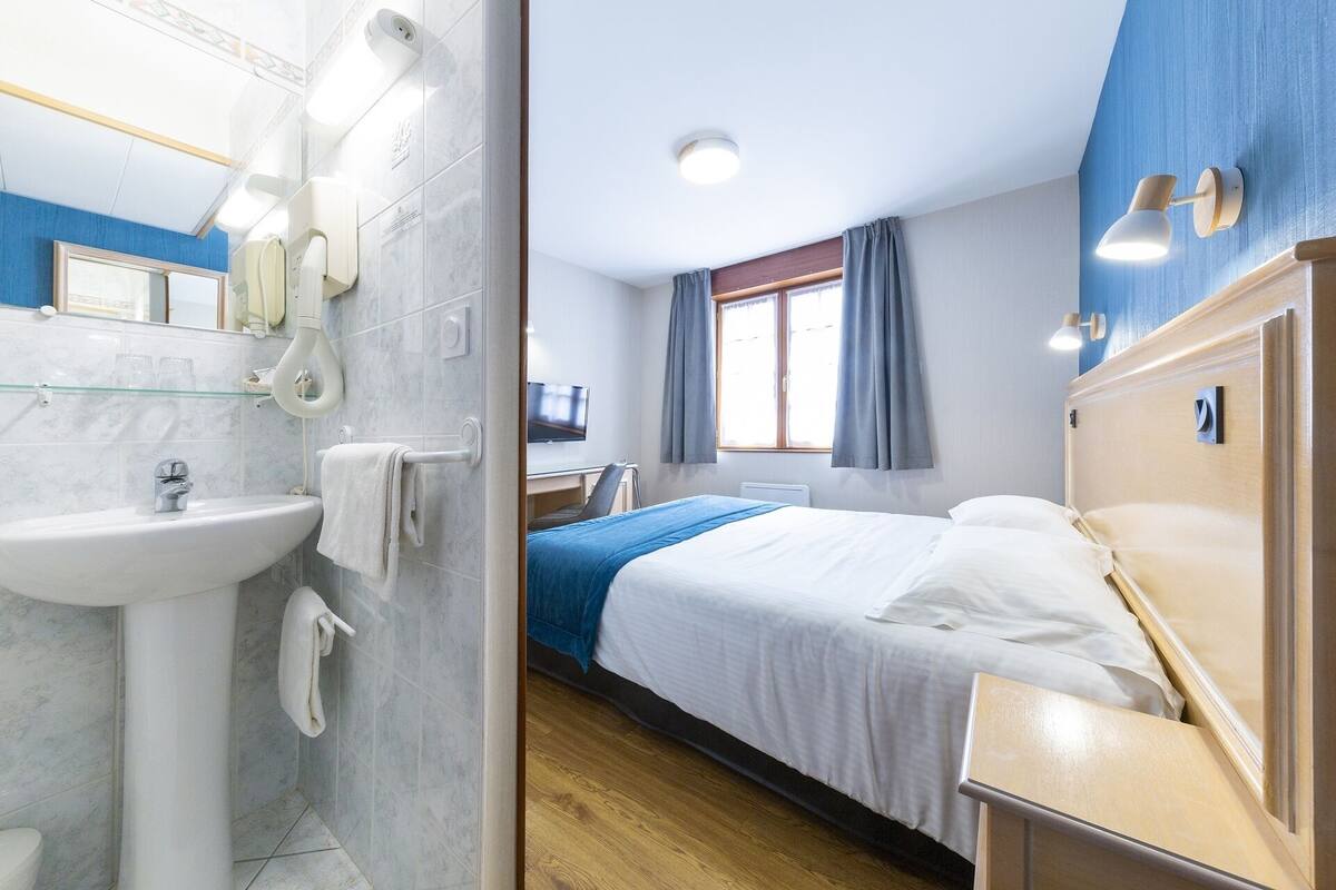 Quarto de hotel com banheiro, cama, mesa de cabeceira e televisão.
