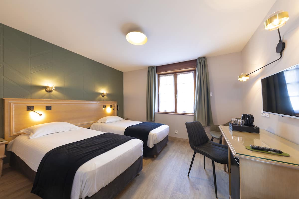 Quarto de hotel com duas camas, uma escrivaninha com cadeira, televisão e janela com cortinas.
