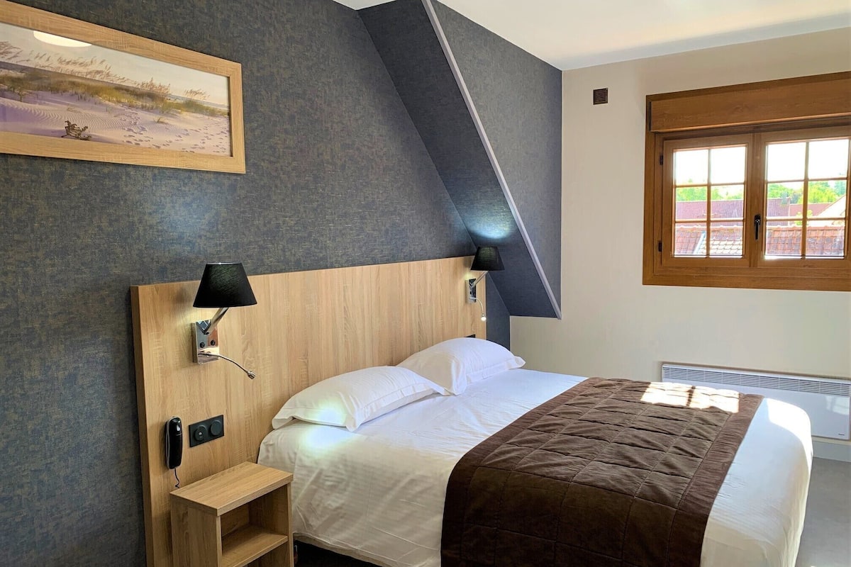 Quarto de hotel com cabeceira de madeira, cama com roupa de cama branca, manta marrom, mesa de cabeceira de madeira pequena, telefone, luminária na parede, pintura e janela com vista para edifícios.
