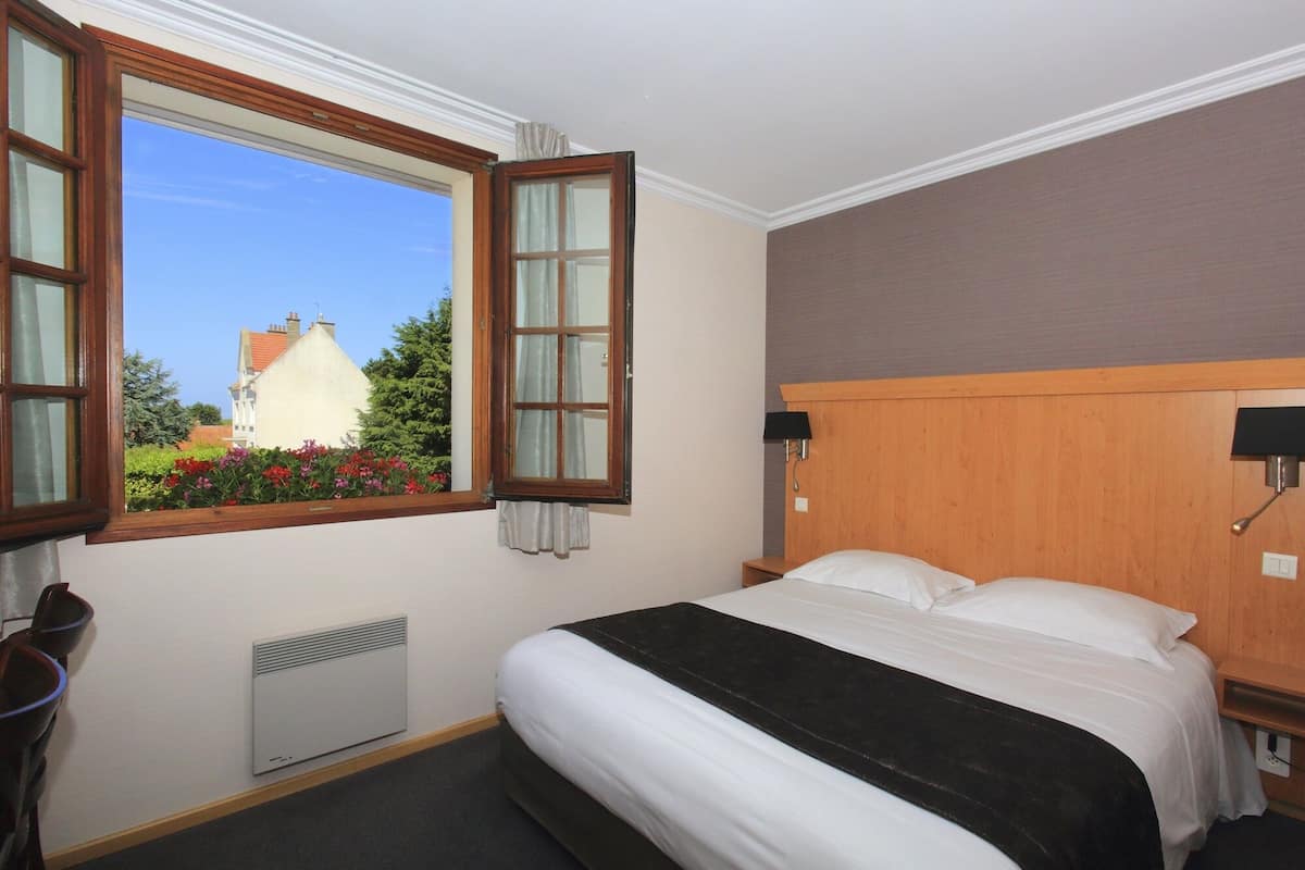 Quarto de hotel com cama, janela com cortinas, aquecedor fixado na parede e vista para uma casa e árvores lá fora.