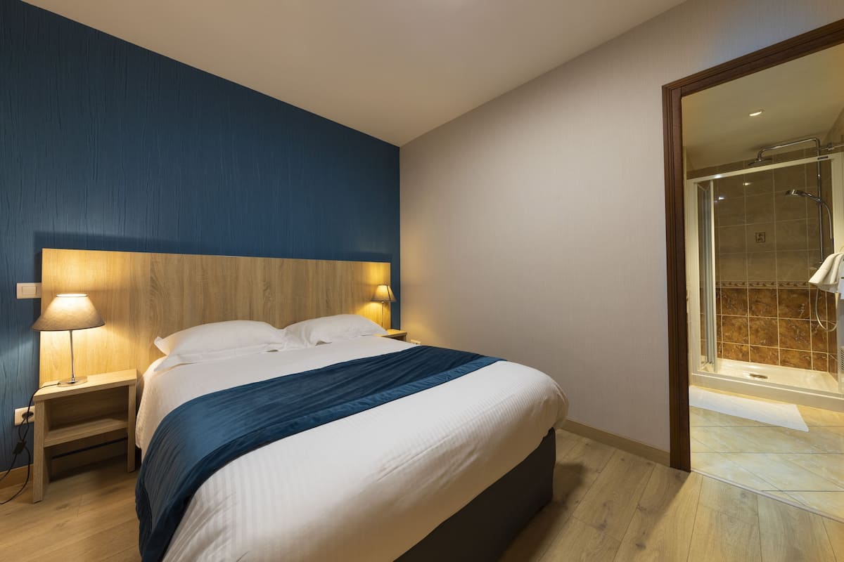 Quarto de hotel com cama, luminárias de cabeceira, painel de cabeceira em madeira, área de chuveiro e piso ladrilhado.