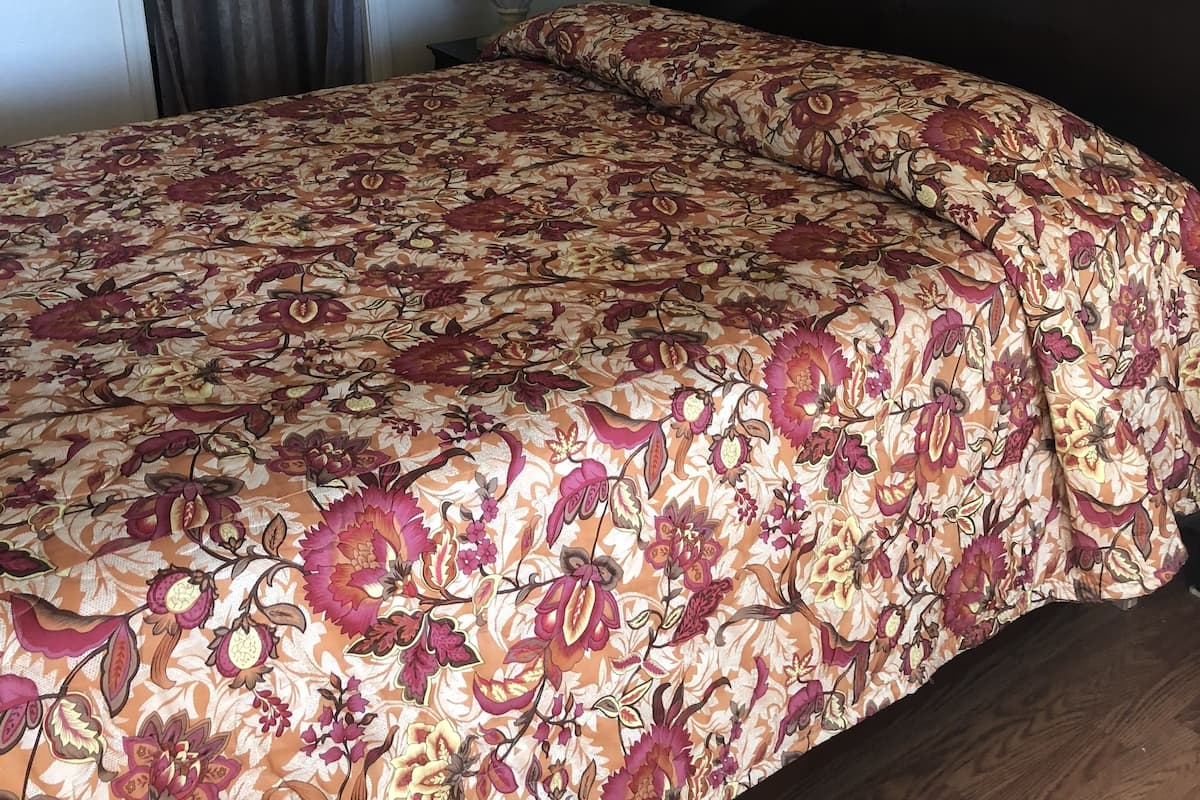 Quarto de hotel com colcha florida, quadro de flores na parede, abajur e janela com cortinas.