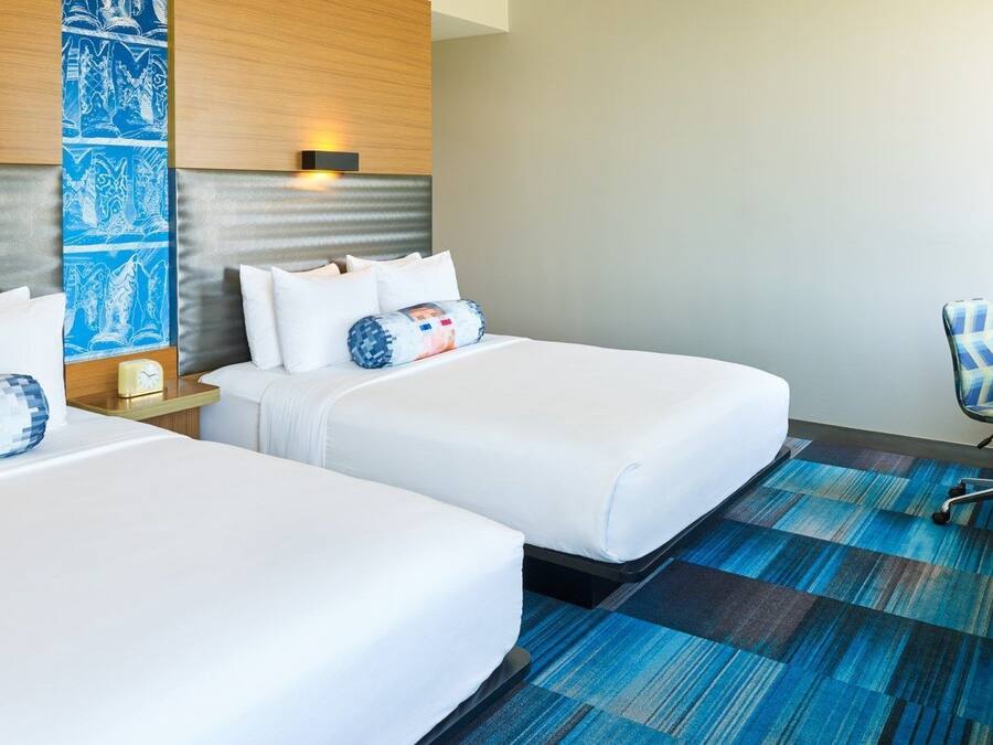 Quarto de hotel com duas camas, uma mesa de cabeceira, uma cadeira e um abajur.