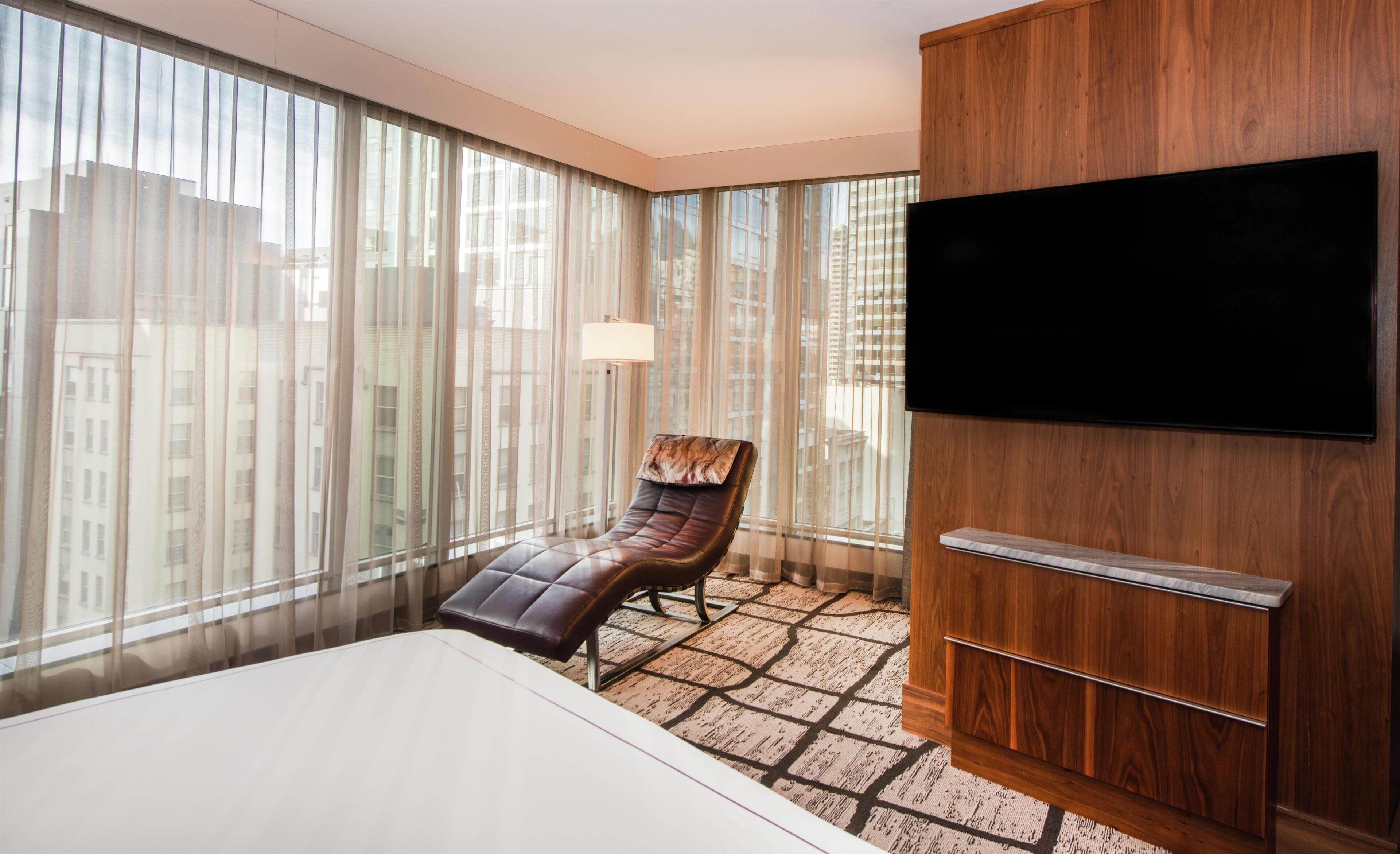 Quarto de hotel com uma cama, uma poltrona de couro, uma TV fixada em um painel de madeira e janelas amplas com cortinas translúcidas.