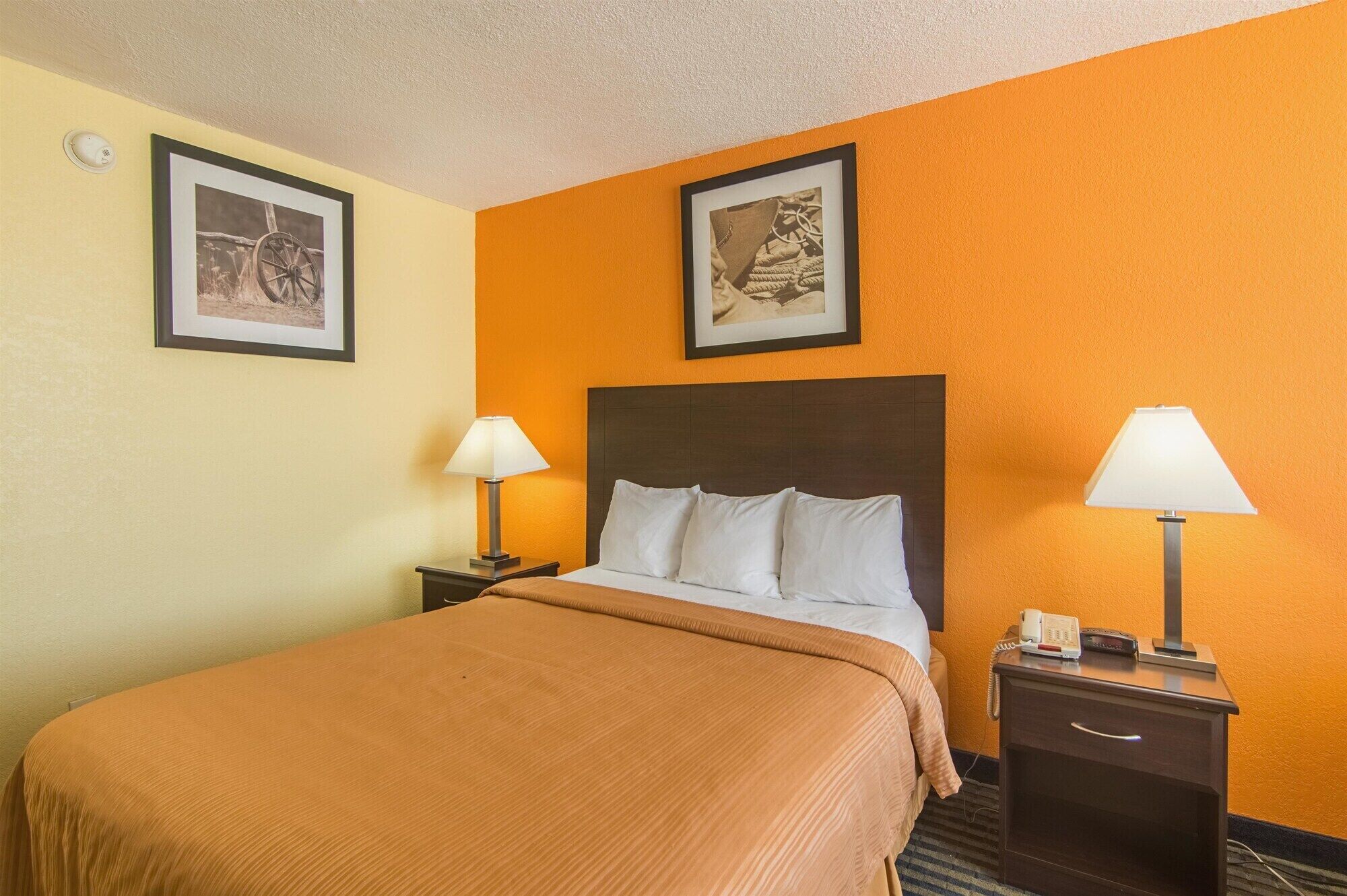 Quarto de hotel com uma cama grande, duas mesas de cabeceira com abajures e duas fotos emolduradas nas paredes.