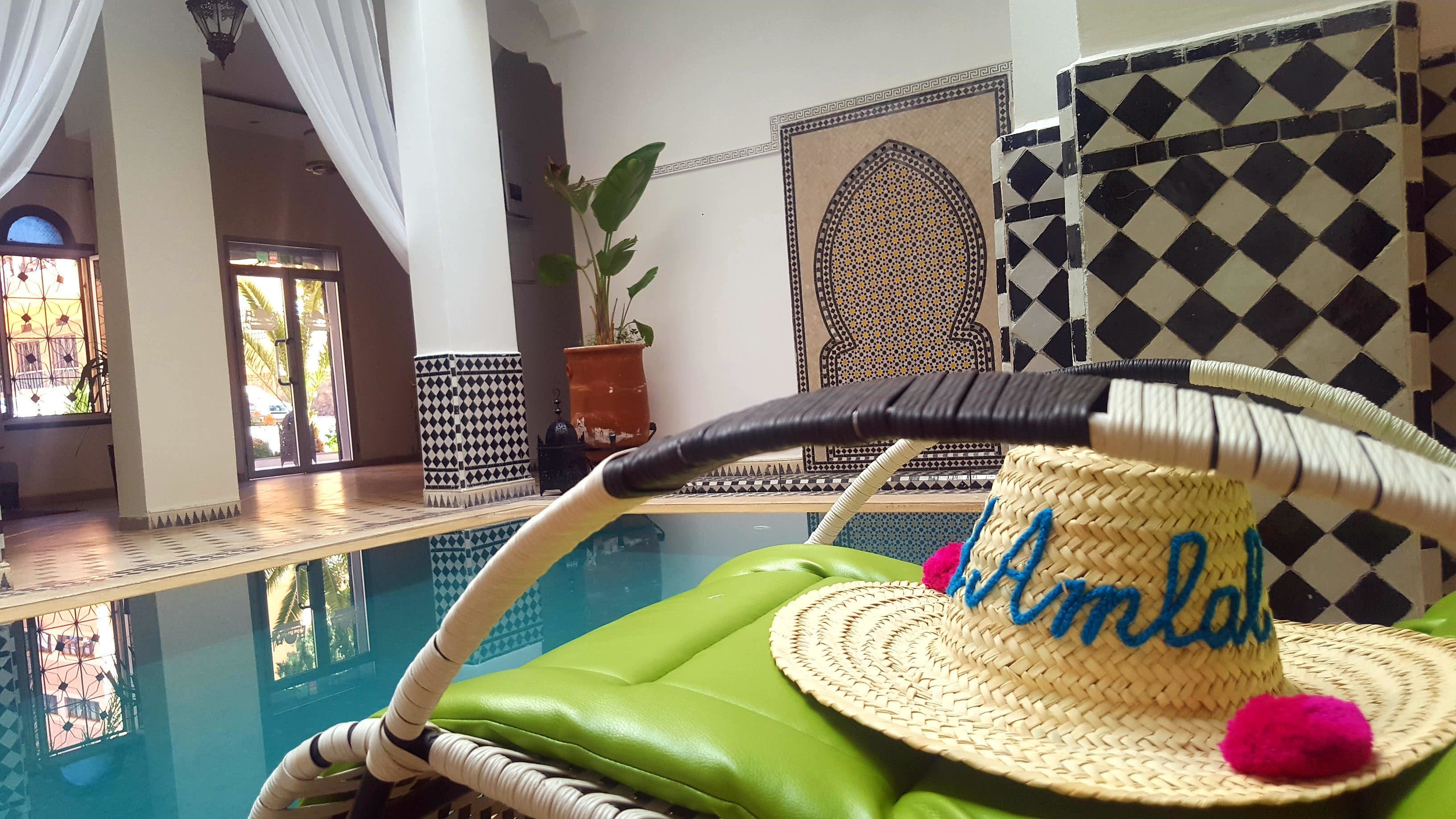 Uma área de piscina com uma espreguiçadeira verde, um chapéu de palha com a inscrição 'Amal' bordada e uma parede decorativa com padrões geométricos.