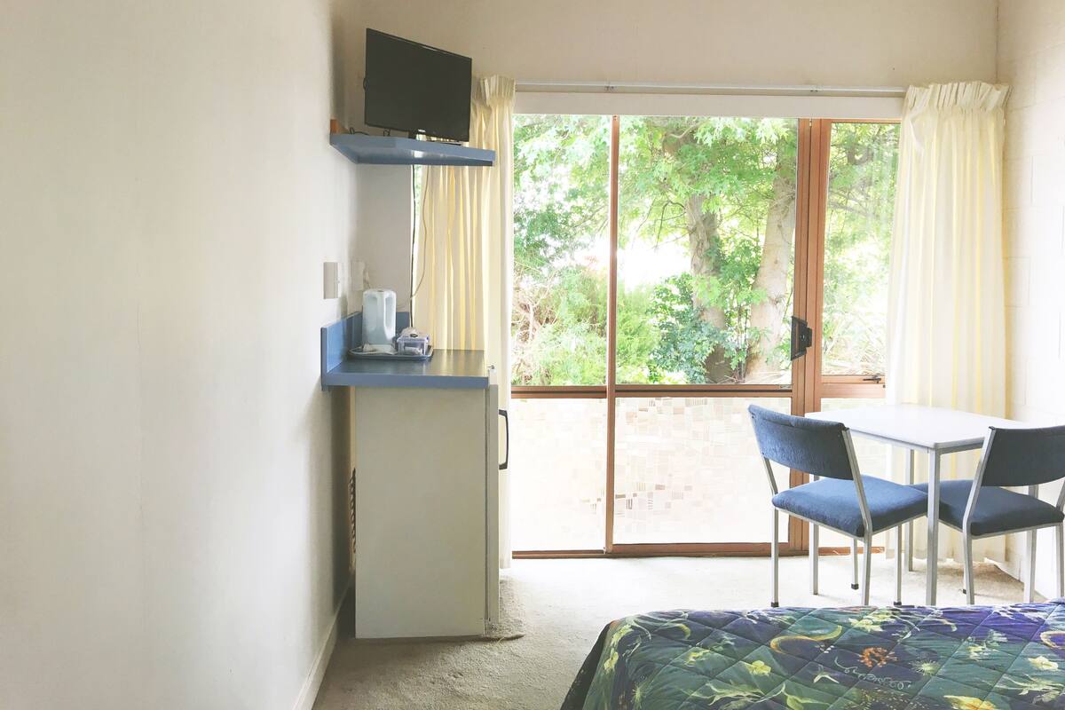 Quarto com cama, escrivaninha com computador, cadeira e televisão em uma estante.