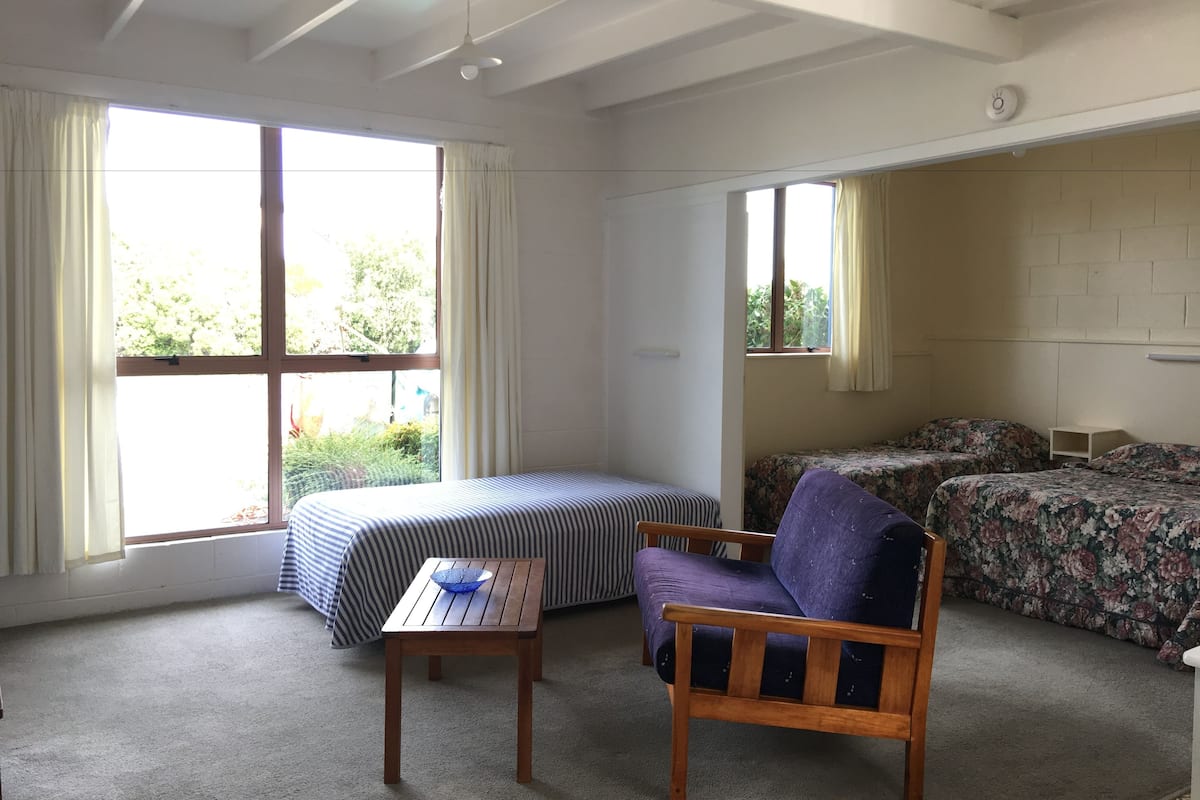 Quarto com cama, uma cadeira, uma mesinha e duas janelas.
