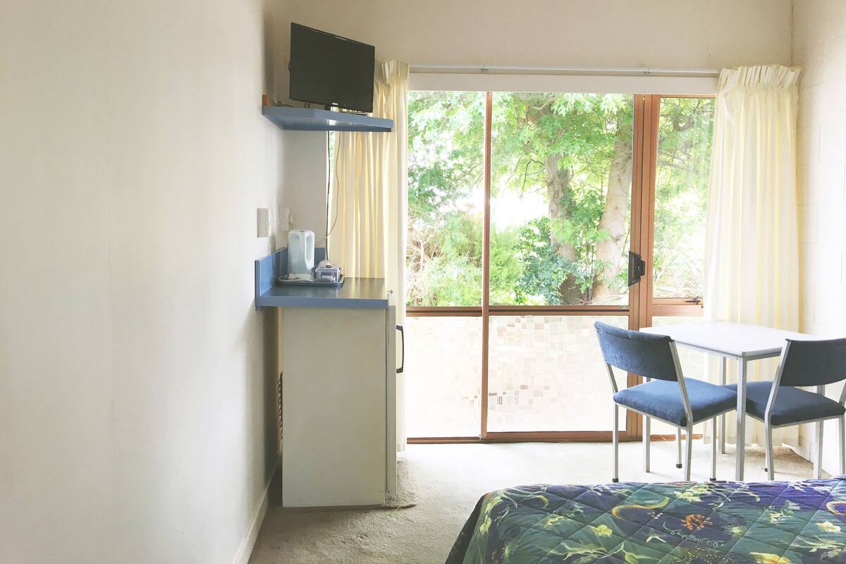 Quarto com cama, escrivaninha com computador, cadeira e televisão em um suporte.