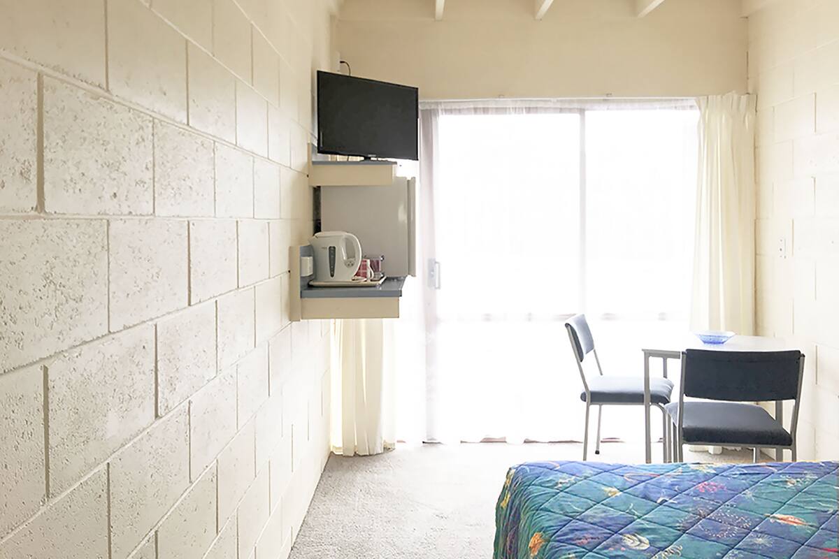 Quarto com cama, escrivaninha, cadeira, televisão fixada na parede e janela com cortinas.