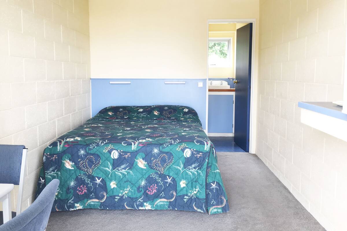 Um pequeno quarto de motel, com uma cama, uma escrivaninha e um banheiro visível por uma porta aberta.