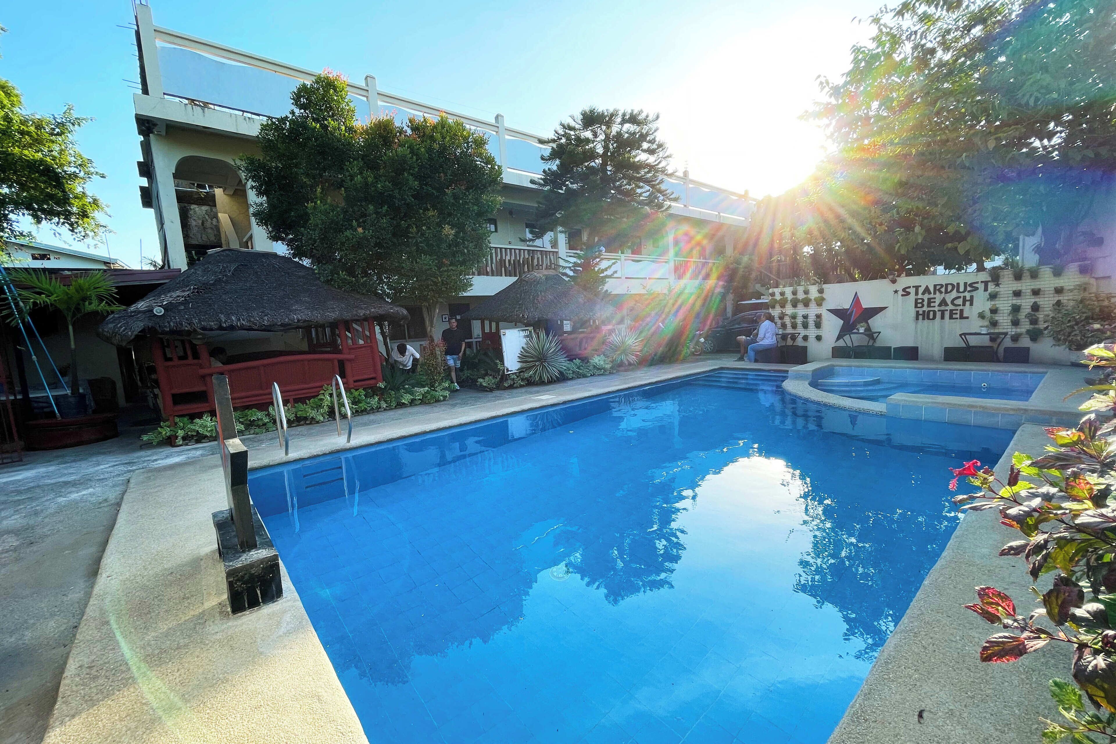Piscina com placa do Stardust Beach Hotel, rodeada por vegetação e um edifício com varanda.