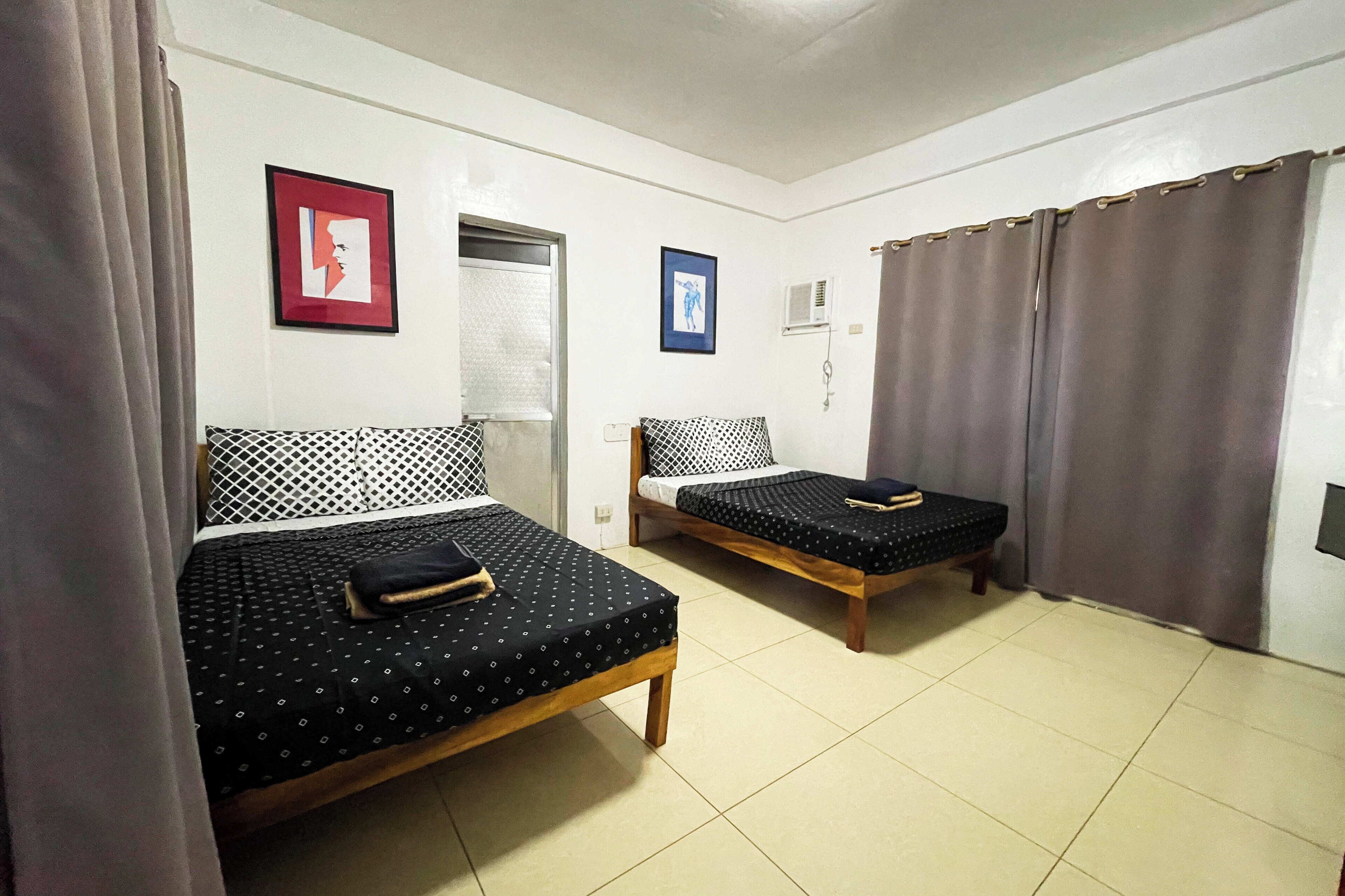 Quarto com duas camas, janela com cortinas, ar-condicionado fixado na parede e um quadro emoldurado.