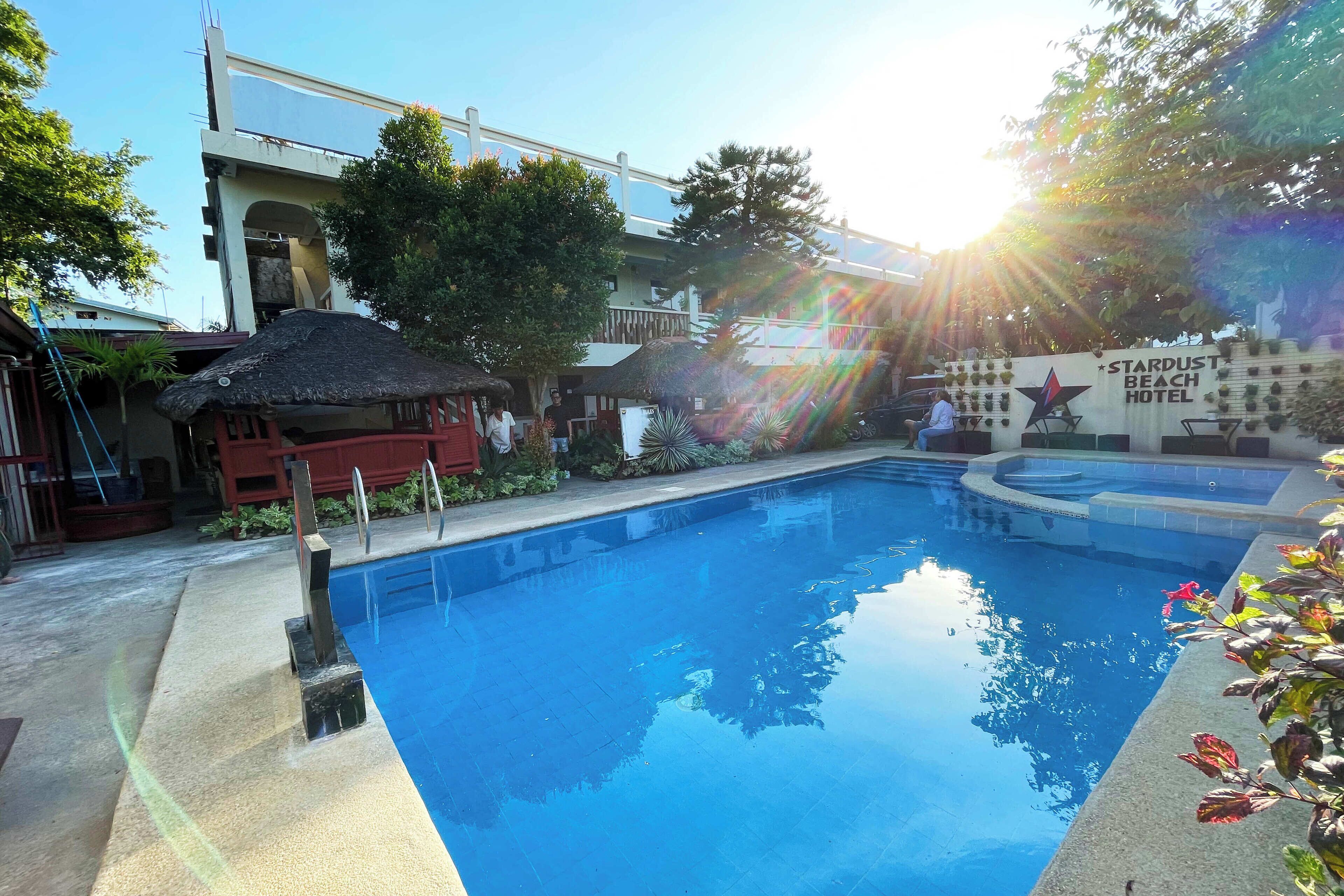 Uma piscina sob um céu ensolarado, um edifício com a inscrição "Stardust Beach Hotel" e uma estrutura coberta de palha.
