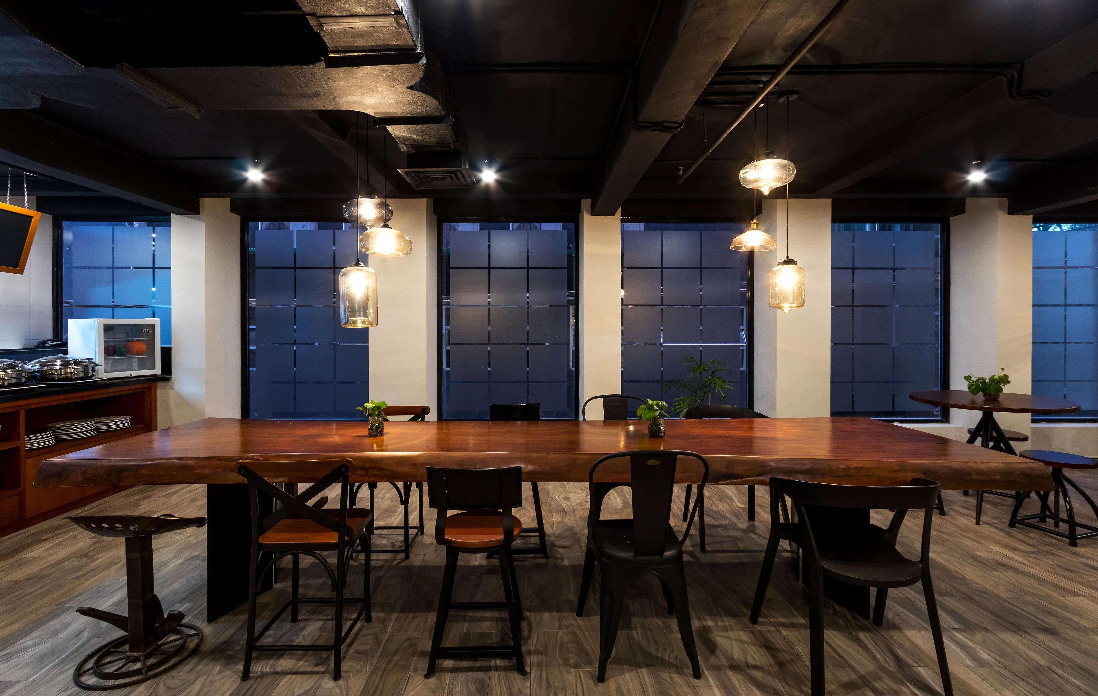 Uma sala de conferência com uma mesa de madeira comprida, cadeiras pretas, luminárias pendentes e janelas amplas com persianas.