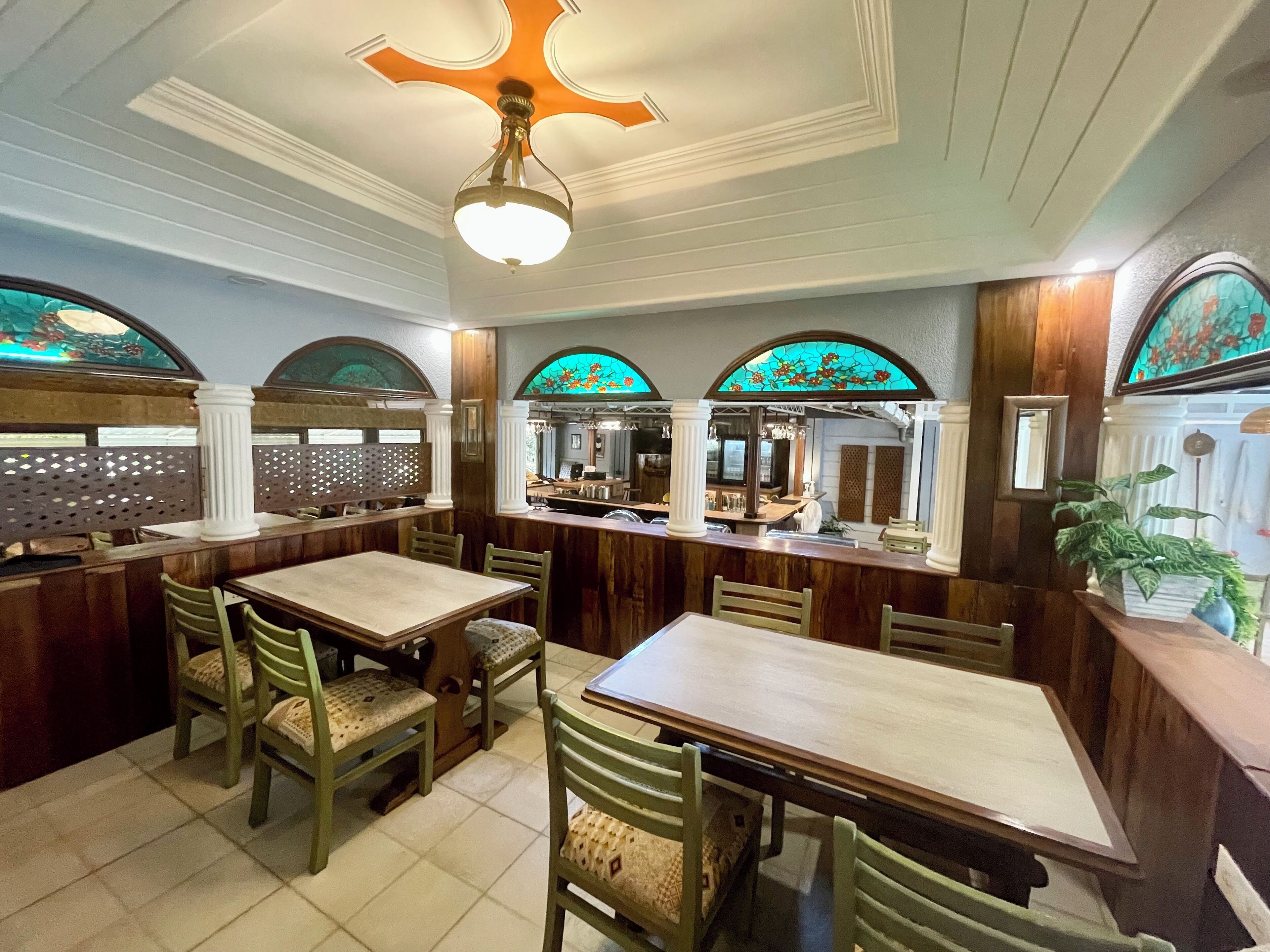 Um restaurante com mesas e cadeiras de madeira, uma ilha central com balcão e janelas decorativas com vitrais.
