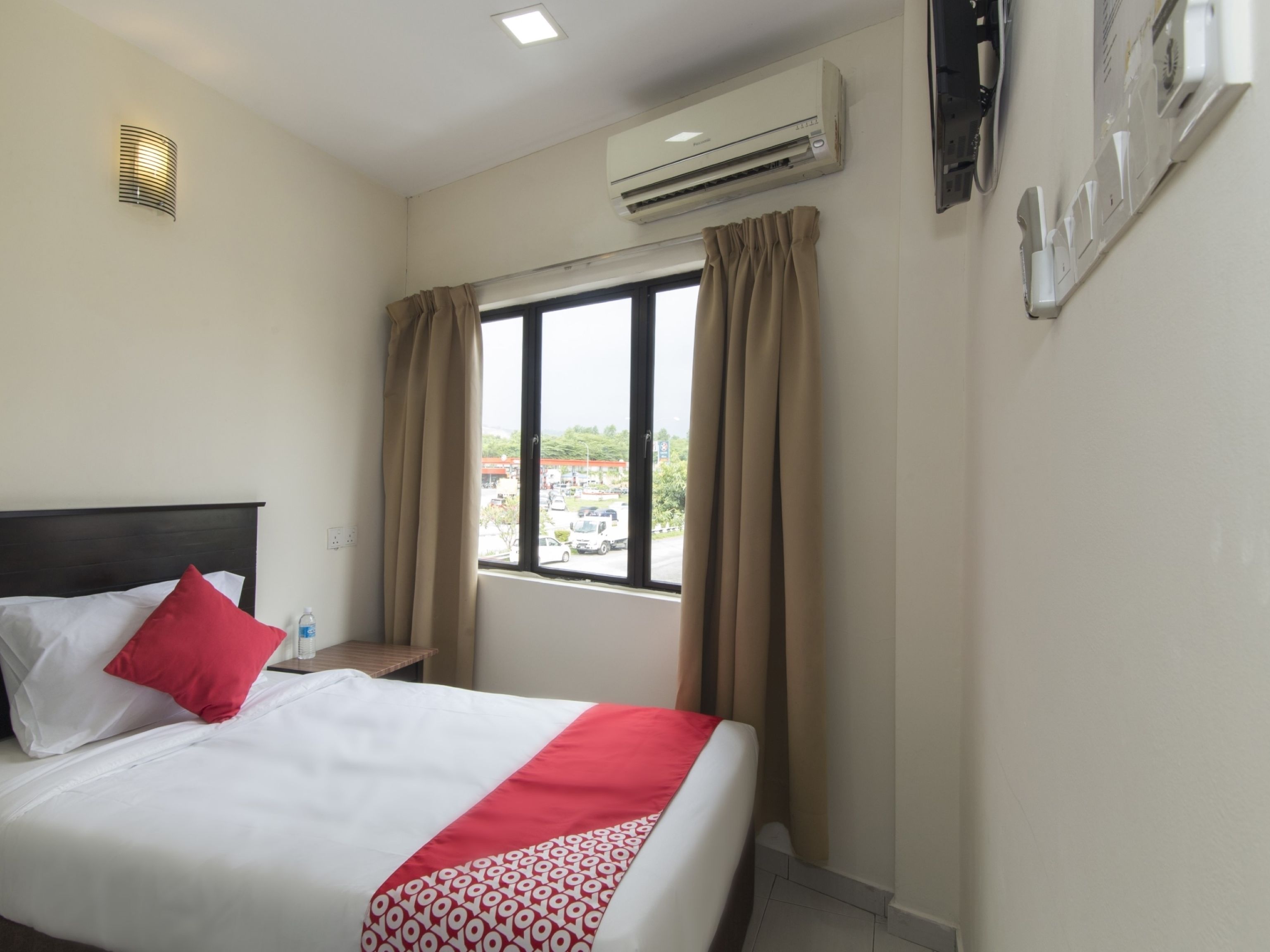 Quarto de hotel com cama, mesa de cabeceira, ar condicionado, janela com cortinas e vista para um estacionamento.