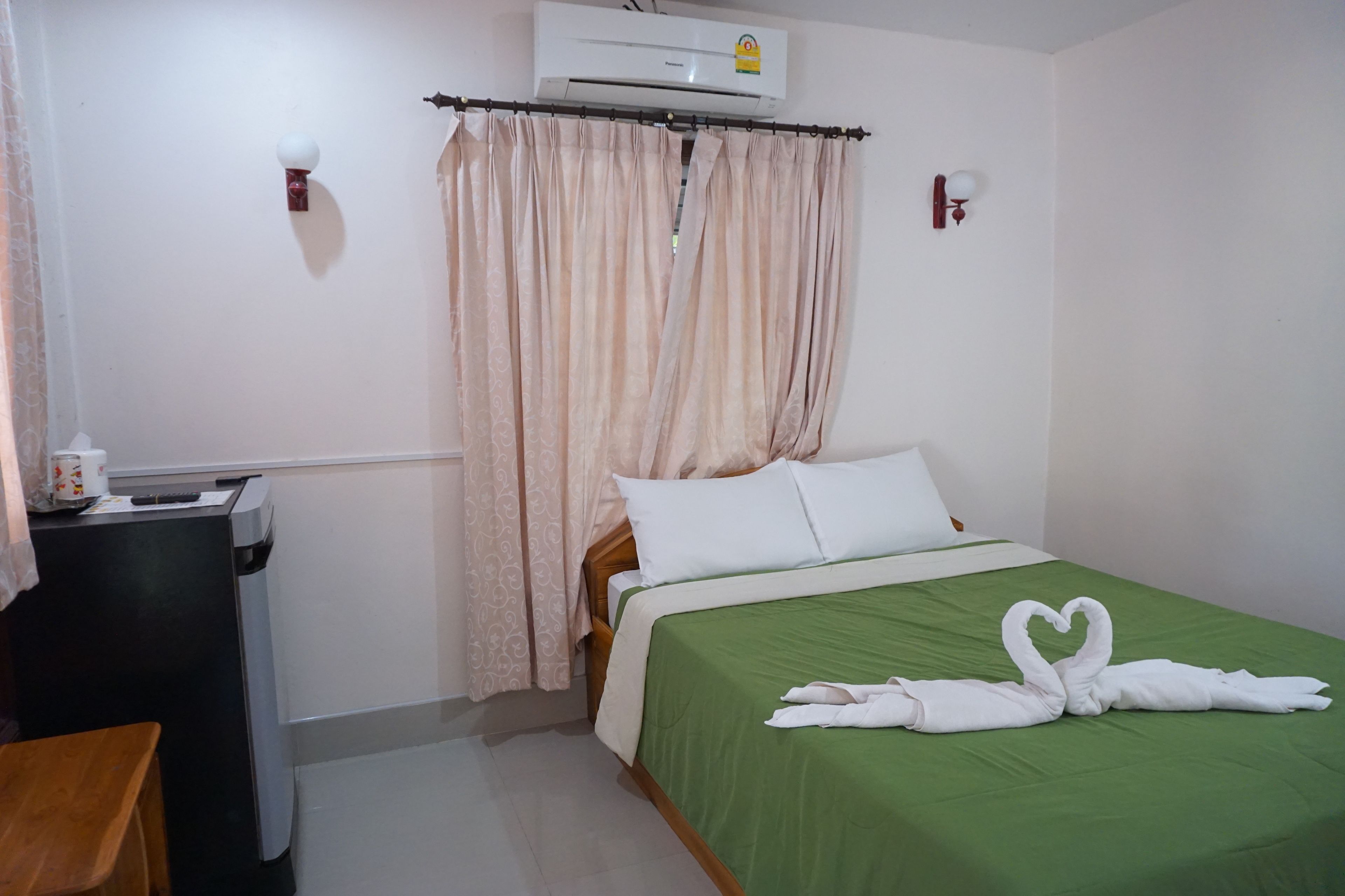 Quarto de hotel com cama verde, almofadas brancas, cortinas bege, mesa de cabeceira de madeira e ar condicionado branco.
