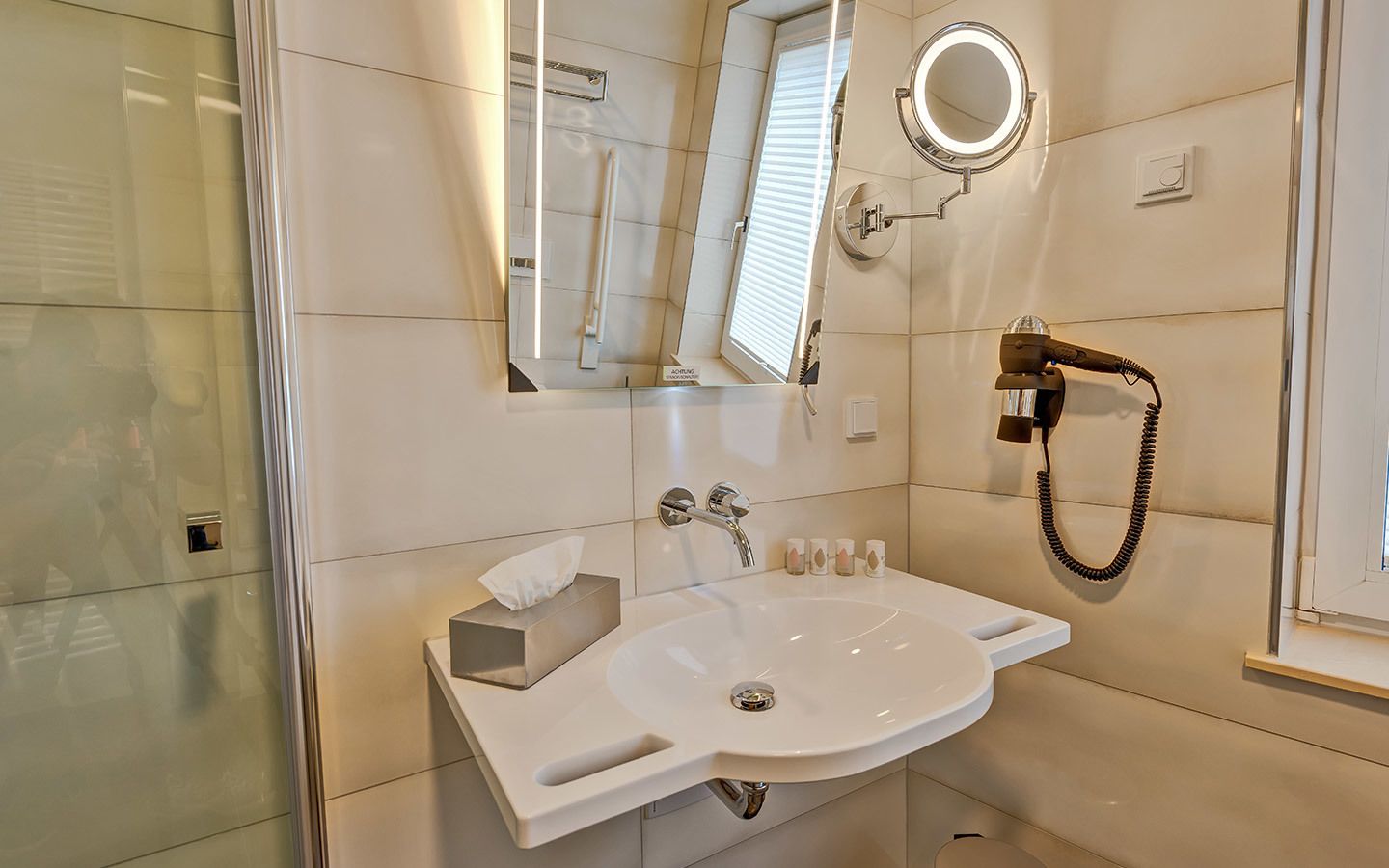 Casa de banho moderna com lavatório branco, espelho, chuveiro manual e secador de cabelo.