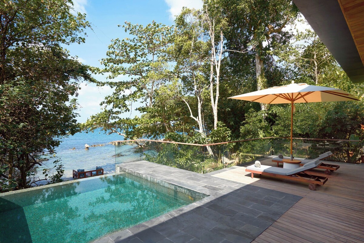 Uma piscina infinita com vista clara para o mar, rodeada por vegetação exuberante e um deque de madeira com espreguiçadeiras e um guarda-sol.