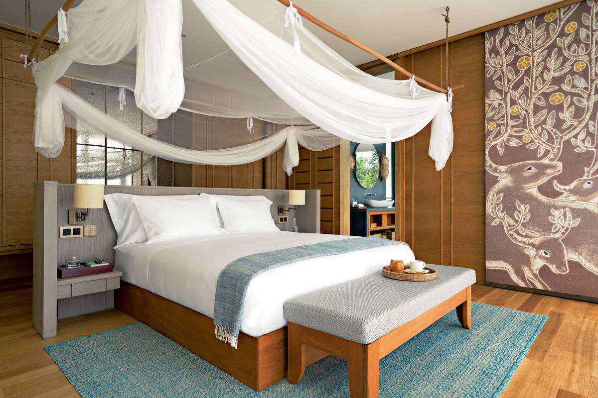 Quarto com cama de dossel, mesas de cabeceira, um banco e um painel decorativo.