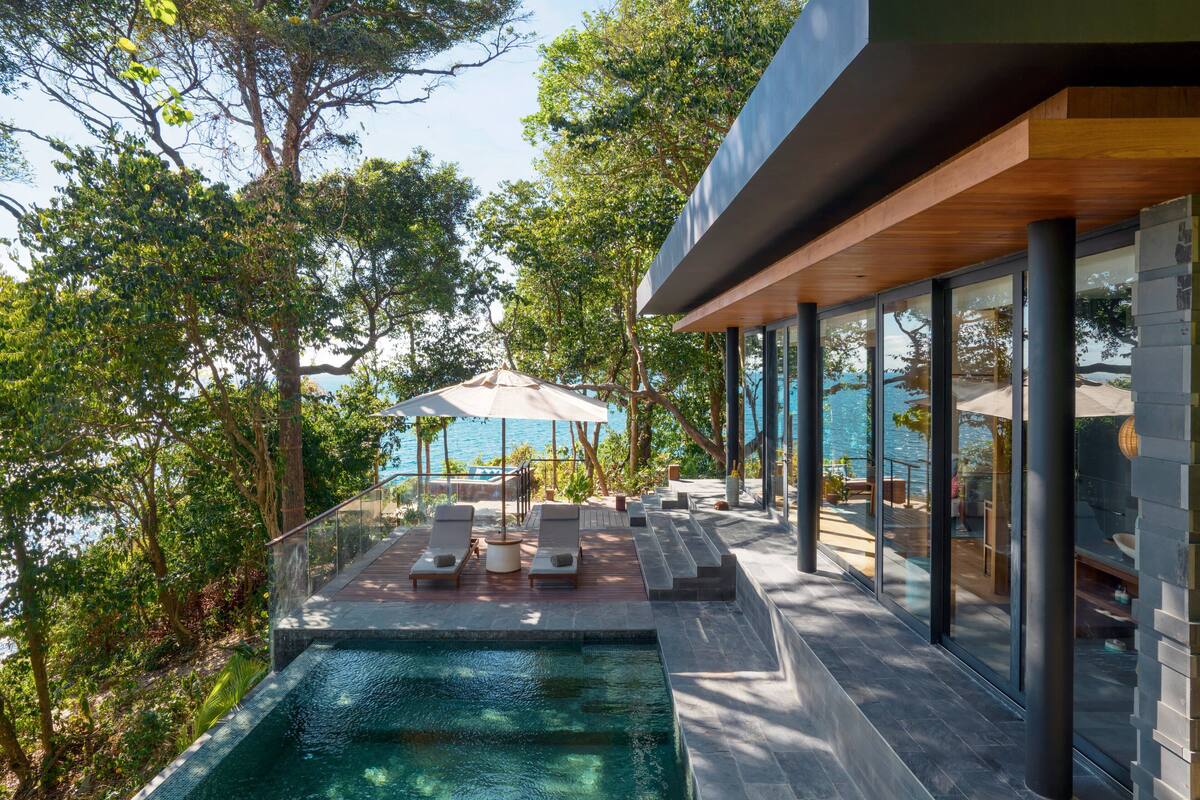 Área moderna de piscina ao ar livre com espreguiçadeiras e guarda-sóis, rodeada por vegetação exuberante e com vista para um corpo de água.