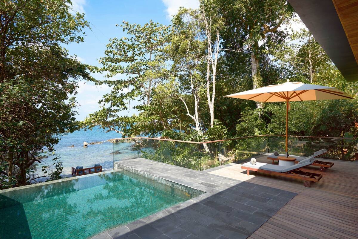 Piscina com deck de madeira e espreguiçadeiras, cercada por árvores e com vista para um corpo de água.