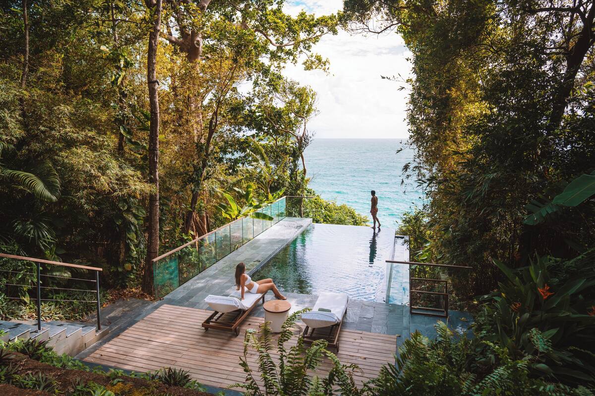 Uma piscina infinita privativa com deck de madeira, rodeada por vegetação exuberante e vista para o mar.