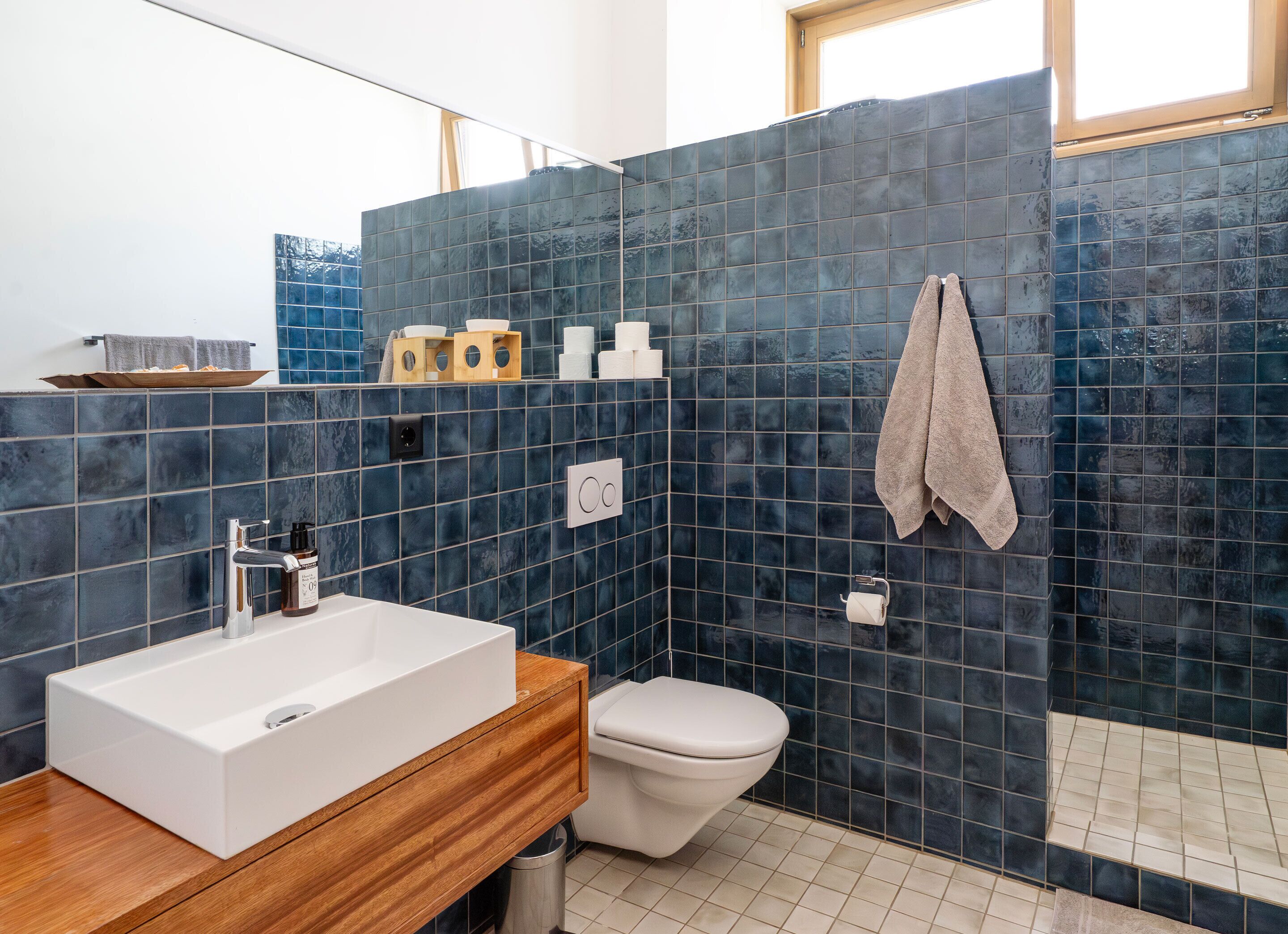 Uma casa de banho moderna com azulejos azul escuro, uma pia branca, toucador de madeira, sanita e uma toalha pendurada na parede.