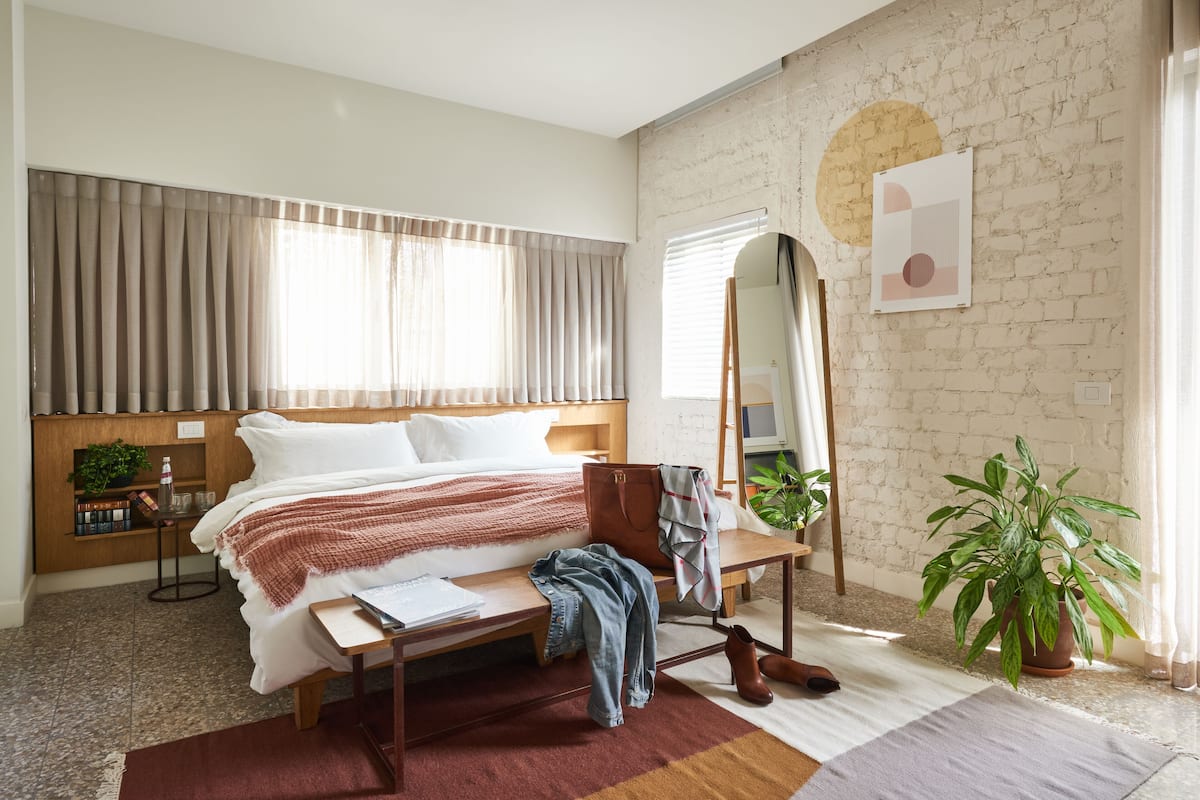 Quarto com cama, mesa de cabeceira, espelho, plantas em vasos e parede de tijolos.