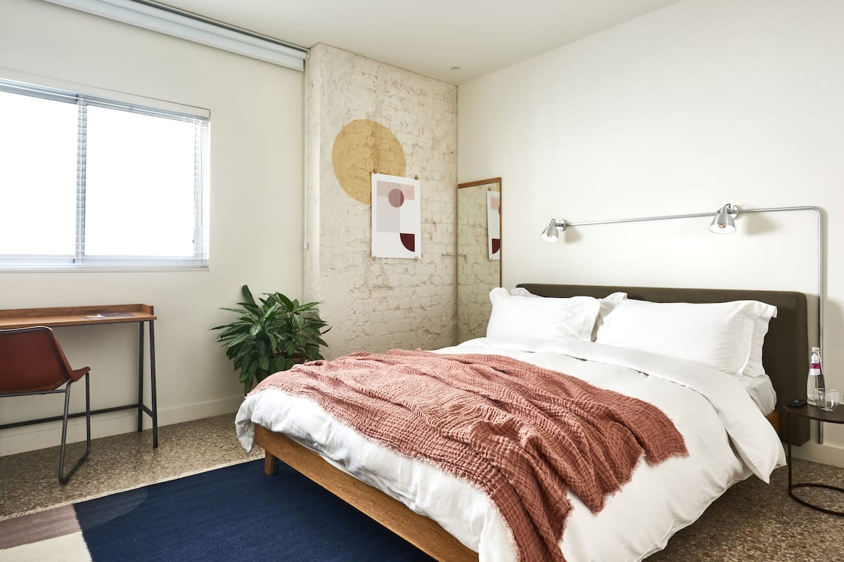 Quarto com cama, escrivaninha, cadeira, planta em vaso e uma parede com uma obra de arte circular.
