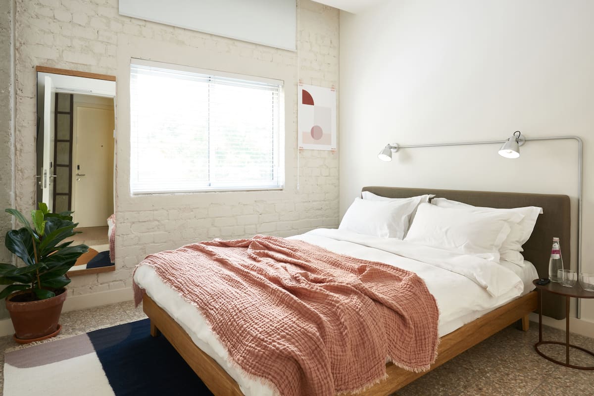 Quarto com cama de madeira, roupa de cama branca, manta rosa, planta em vaso, espelho, janela com persianas e luminária fixada na parede.