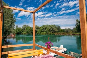Um deck de madeira com um banco e uma mesa, com vista para um rio calmo, barcos e vegetação ao fundo.