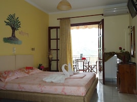 Quarto de hotel com cama, janela com cortinas, varanda com mesa e cadeiras, e parede com ilustração de árvore.