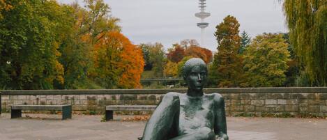 Estátua de uma figura sentada em um parque, com árvores e uma torre ao fundo.