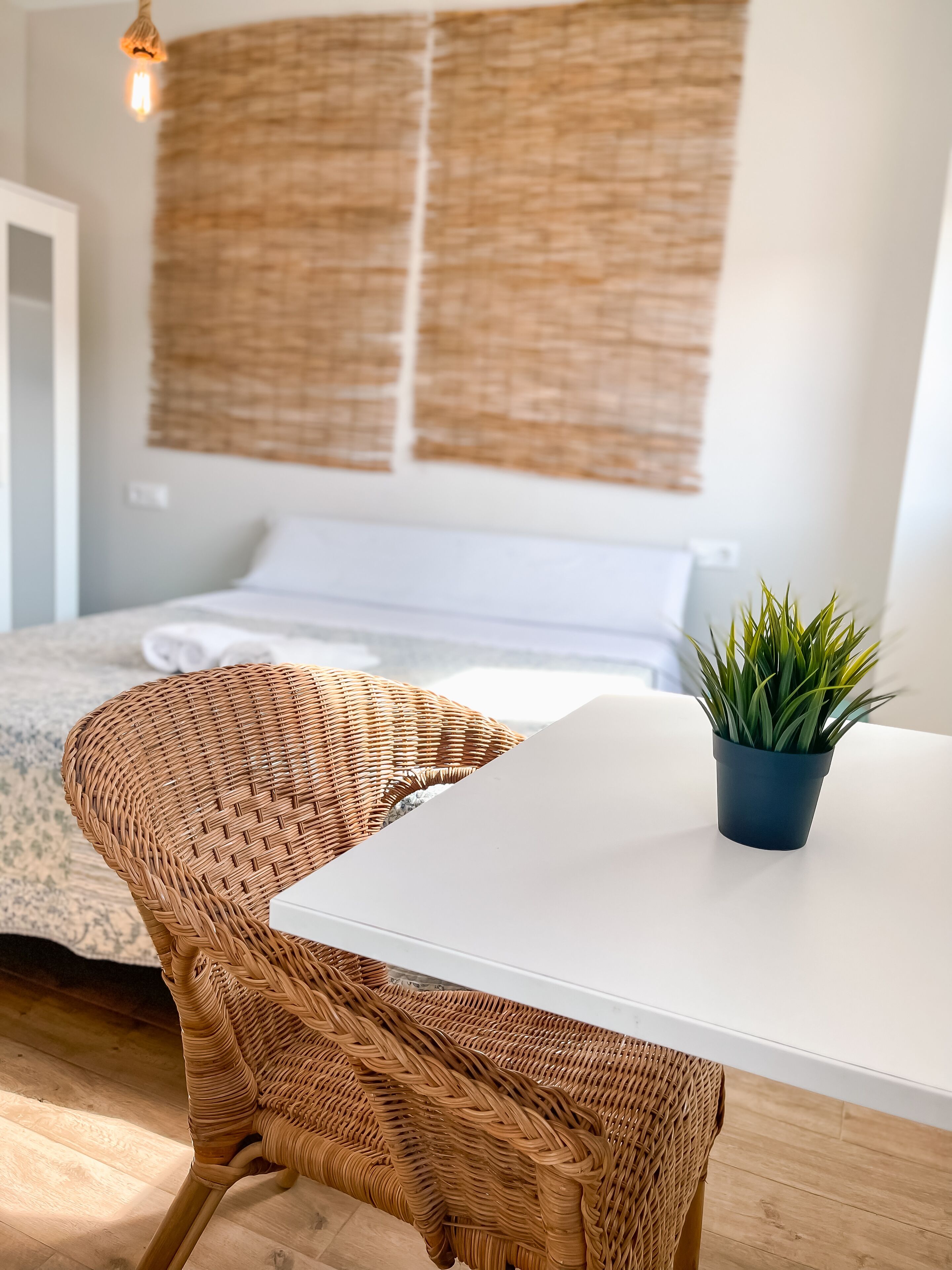 Quarto com cama, cadeira de vime, mesa branca e planta em vaso.