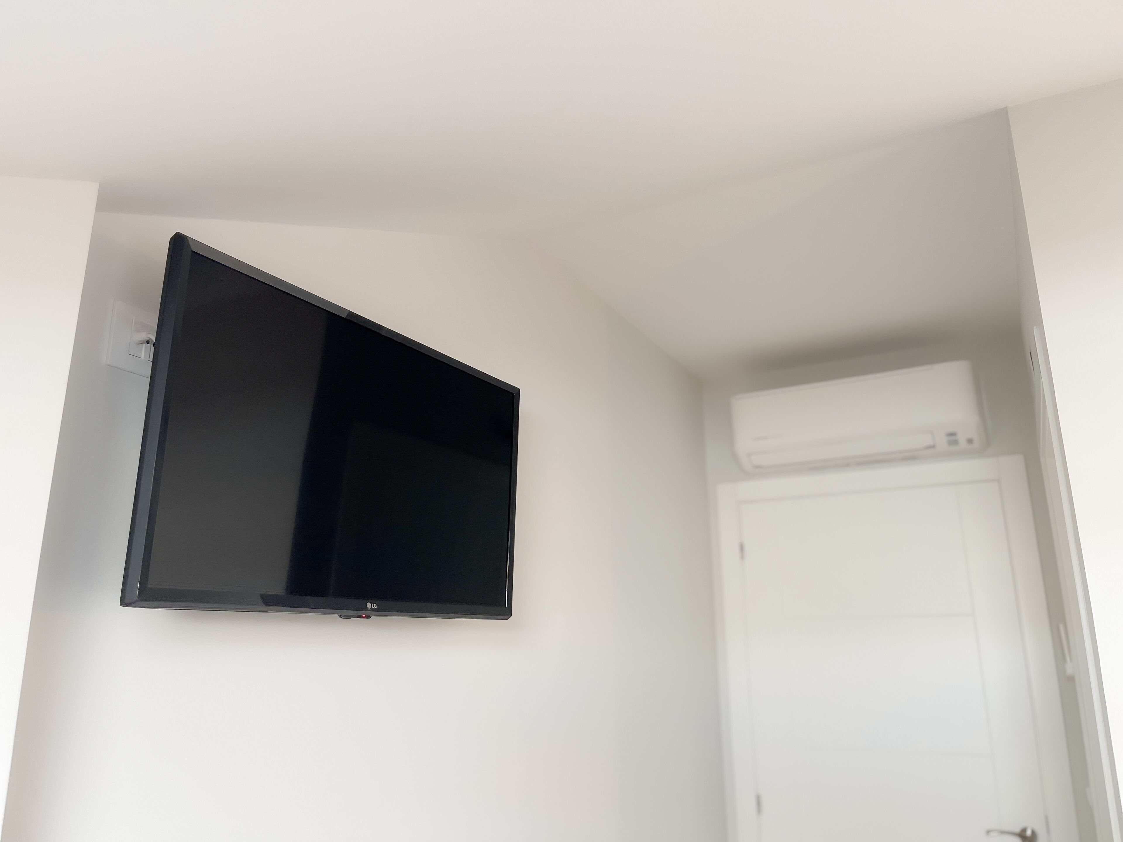 Quarto com televisão de tela plana fixada na parede, porta branca e unidade de ar condicionado acima da porta.