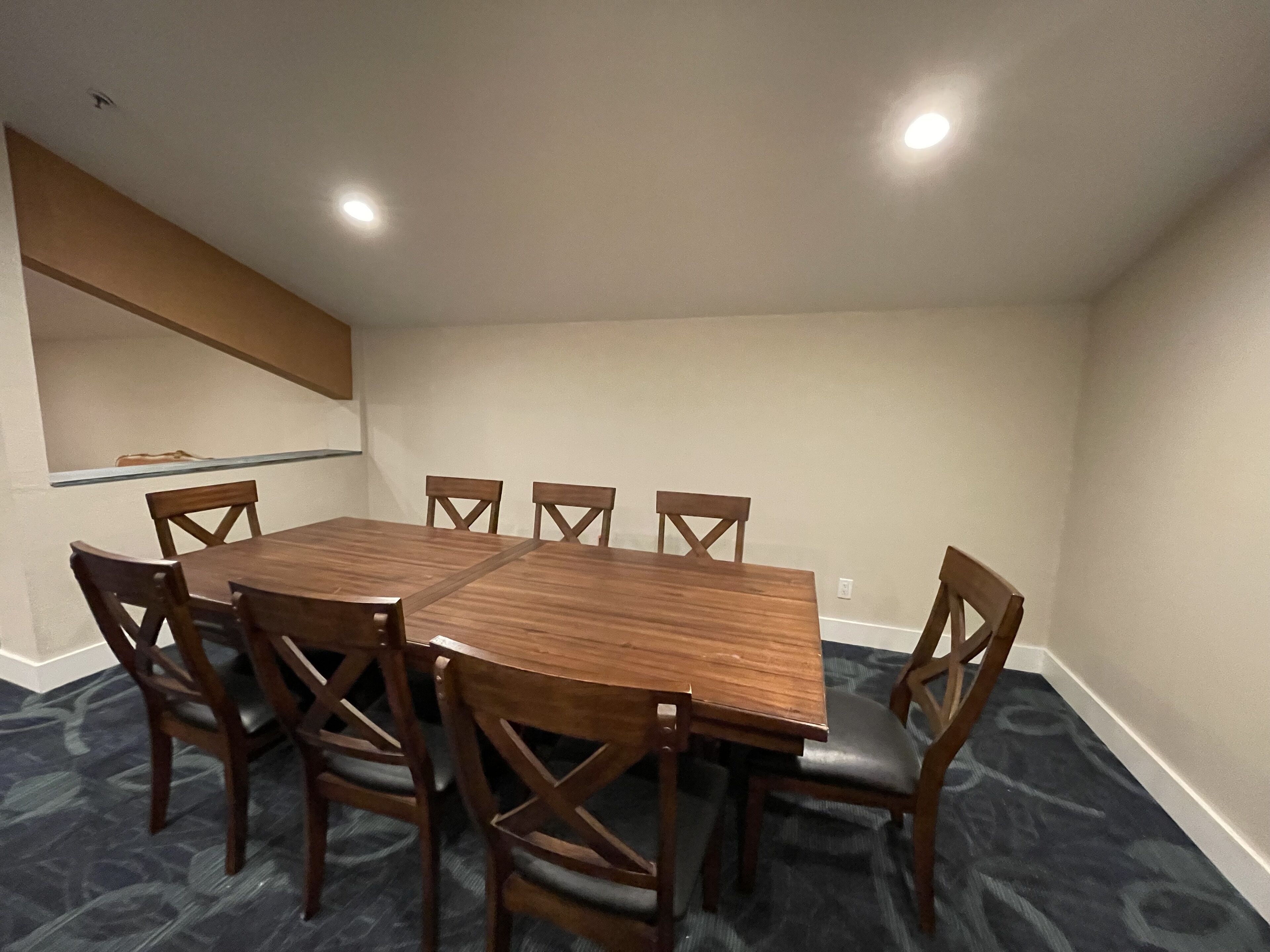 Uma sala de conferências com uma mesa de madeira retangular rodeada por seis cadeiras, piso coberto de carpete e uma parede branca com luminária embutida no teto.