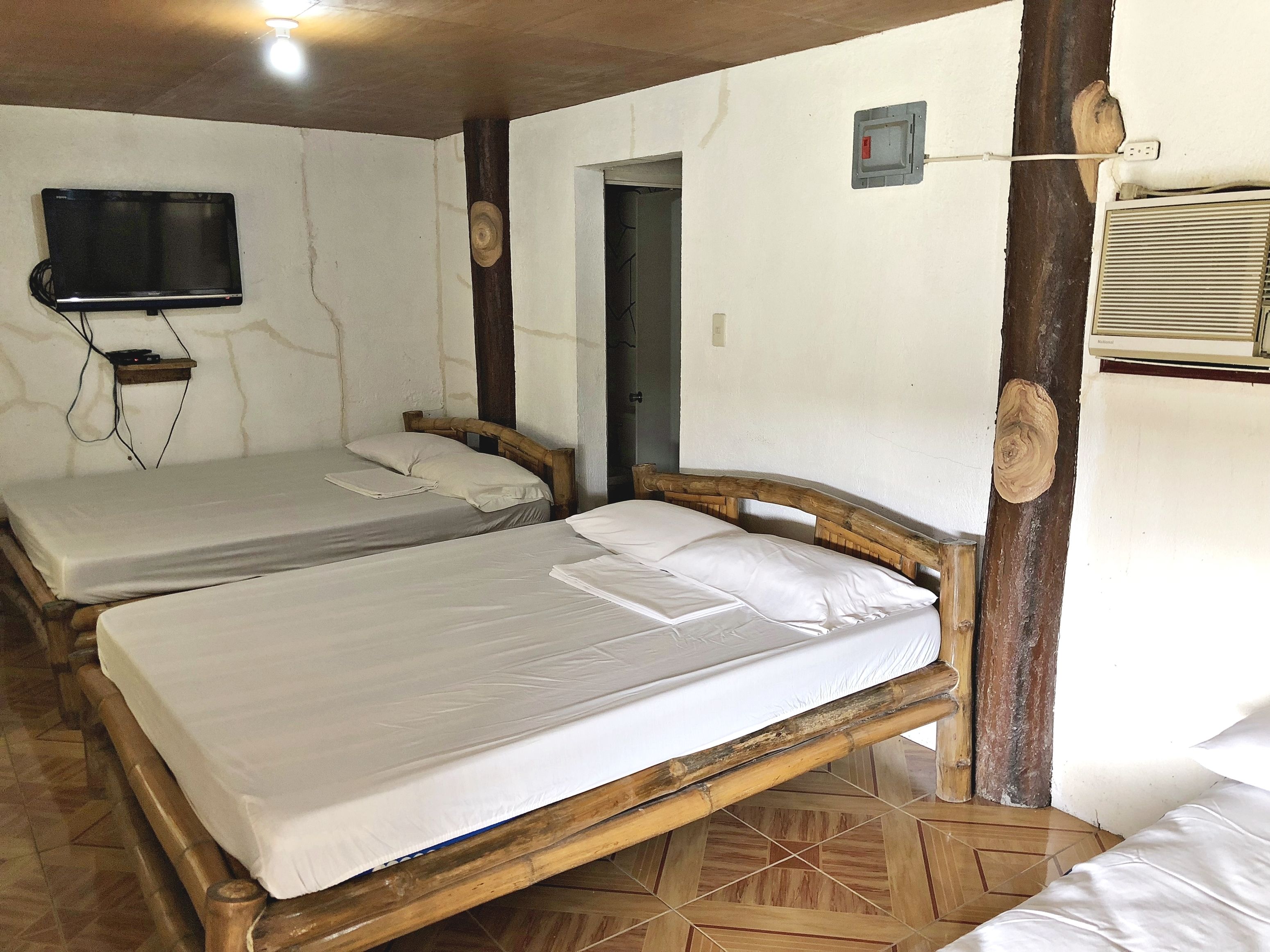 Um quarto com duas camas de madeira, uma televisão fixada na parede e uma porta que dá para outro quarto.