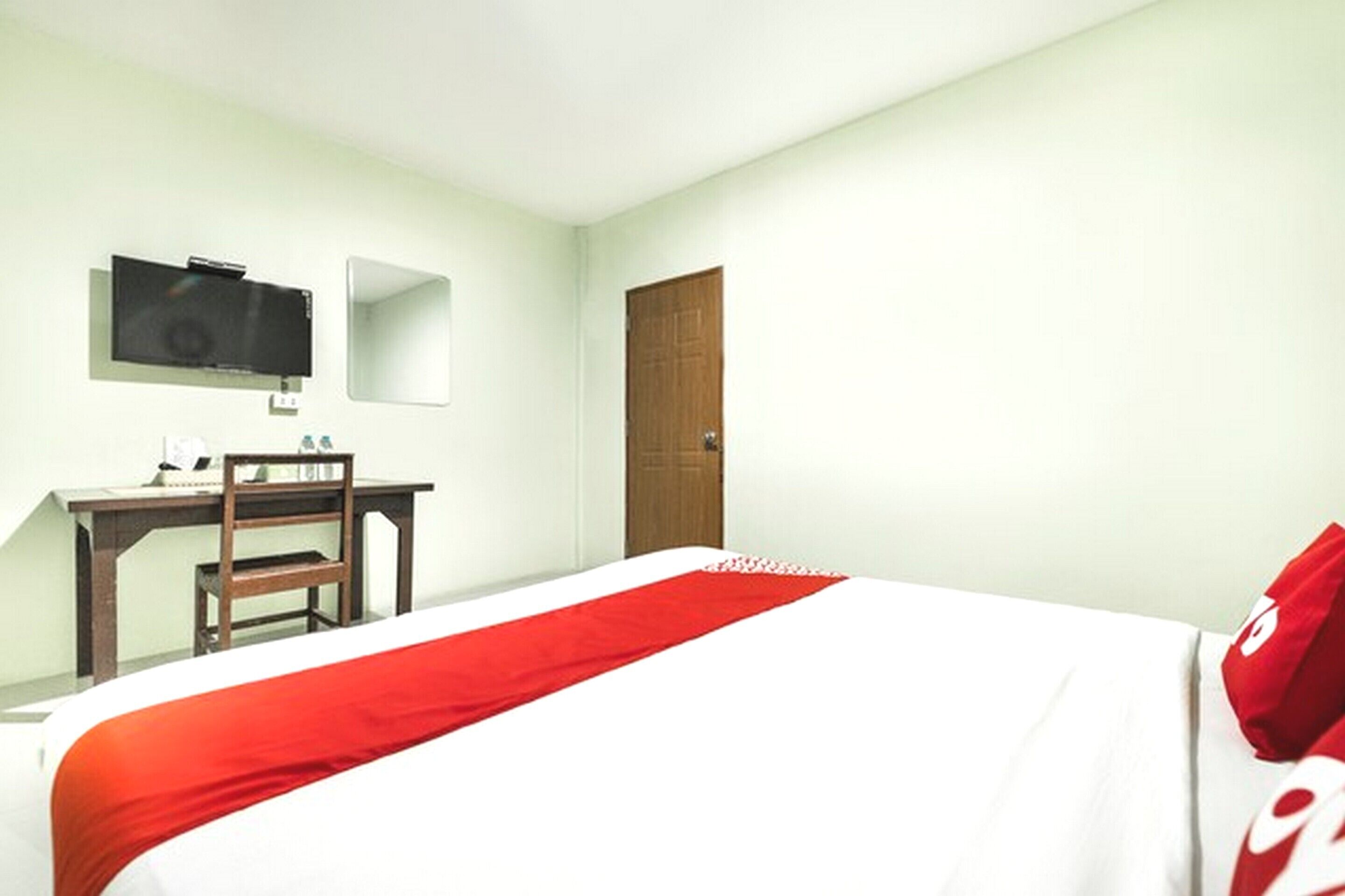 Quarto de hotel com uma cama, uma escrivaninha, uma cadeira, uma televisão presa à parede e uma porta de madeira.