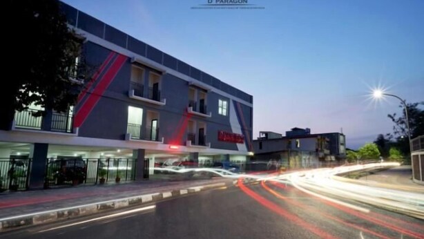 Um edifício moderno com uma placa 'D PARAGON' em destaque, uma área de estacionamento coberta e uma estrada curva com rastros de luz que indicam o tráfego.