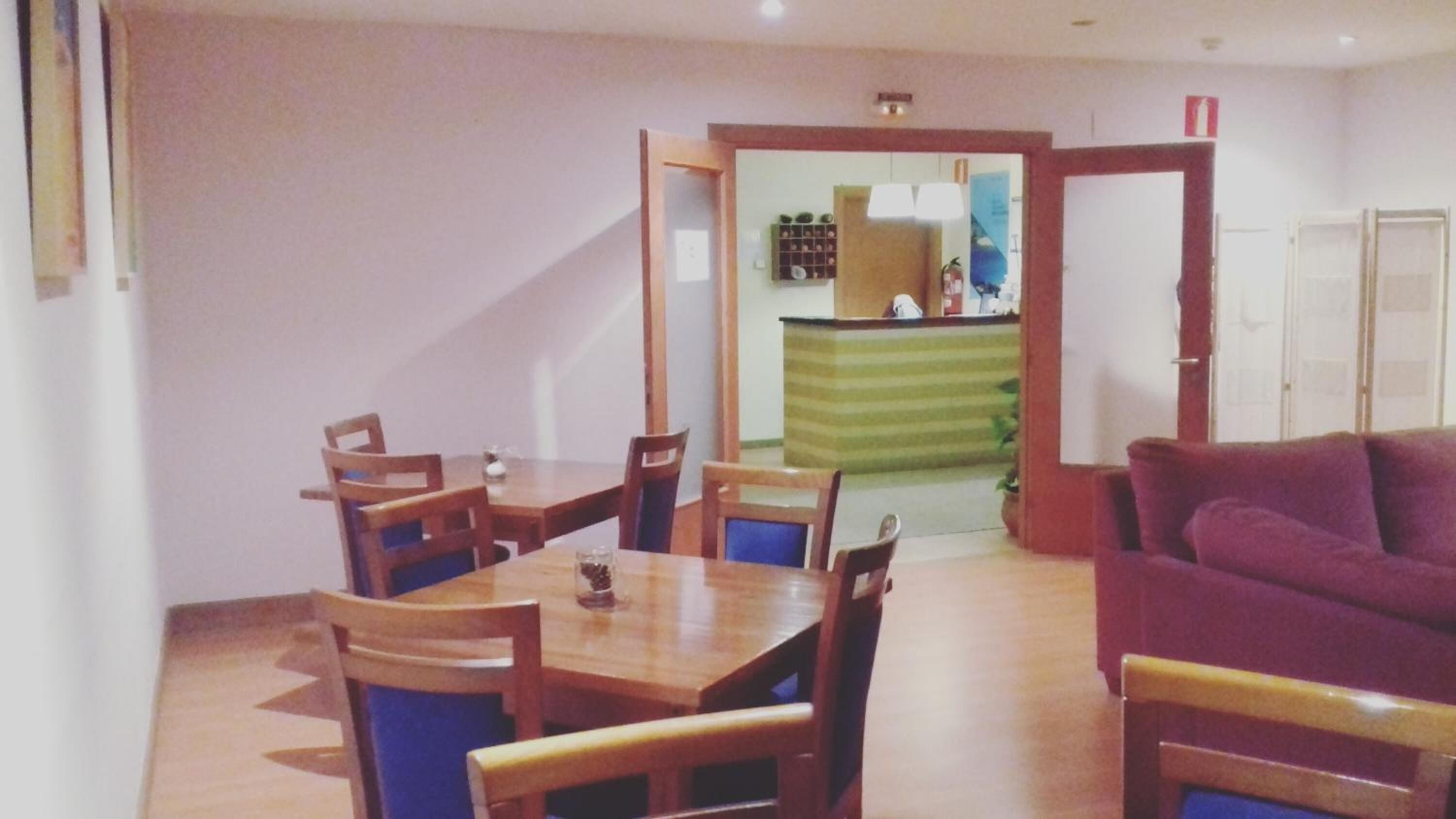 Uma área de refeições com mesa e cadeiras de madeira, um sofá roxo e uma cozinha ao fundo.