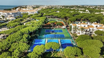 Vista aérea de uma área residencial com quadra de tênis, cercada por vegetação exuberante e edifícios brancos com telhados laranja.