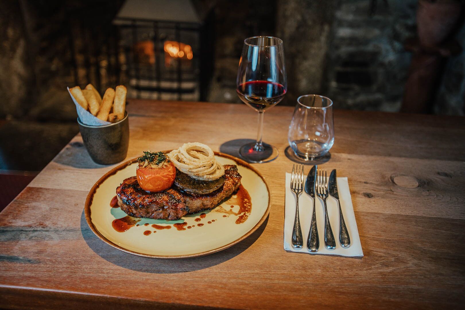 Bife com batatas fritas e anéis de cebola em um prato, acompanhado por um copo de vinho tinto e um copo de água, com talheres e guardanapo sobre uma mesa de madeira.
