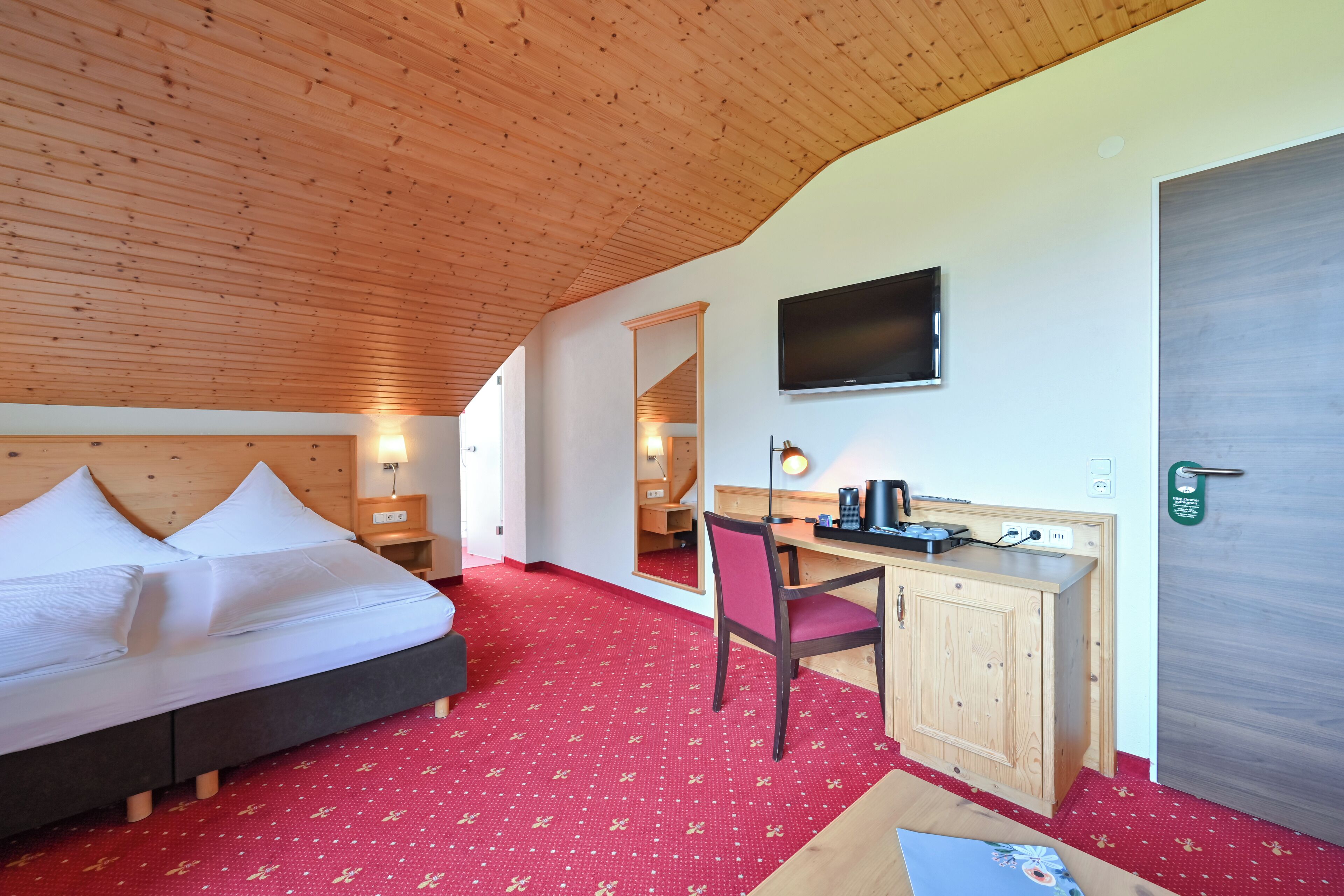 Quarto de hotel com teto de madeira, cama com roupa de cama branca, escrivaninha com cadeira, televisão e porta com puxador.